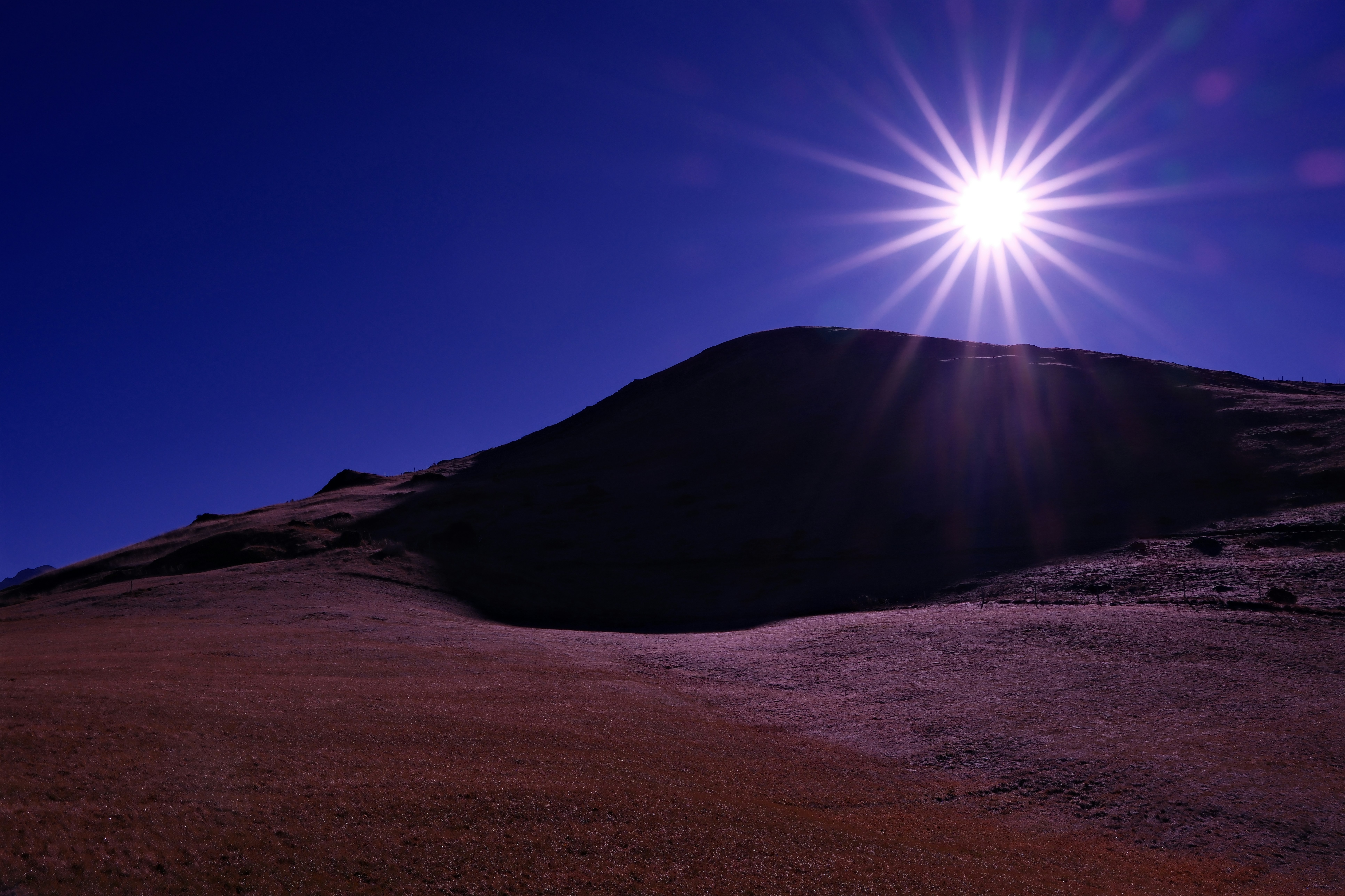 Bright sun radiating over a rugged hillside, casting dramatic shadows on the textured landscape.