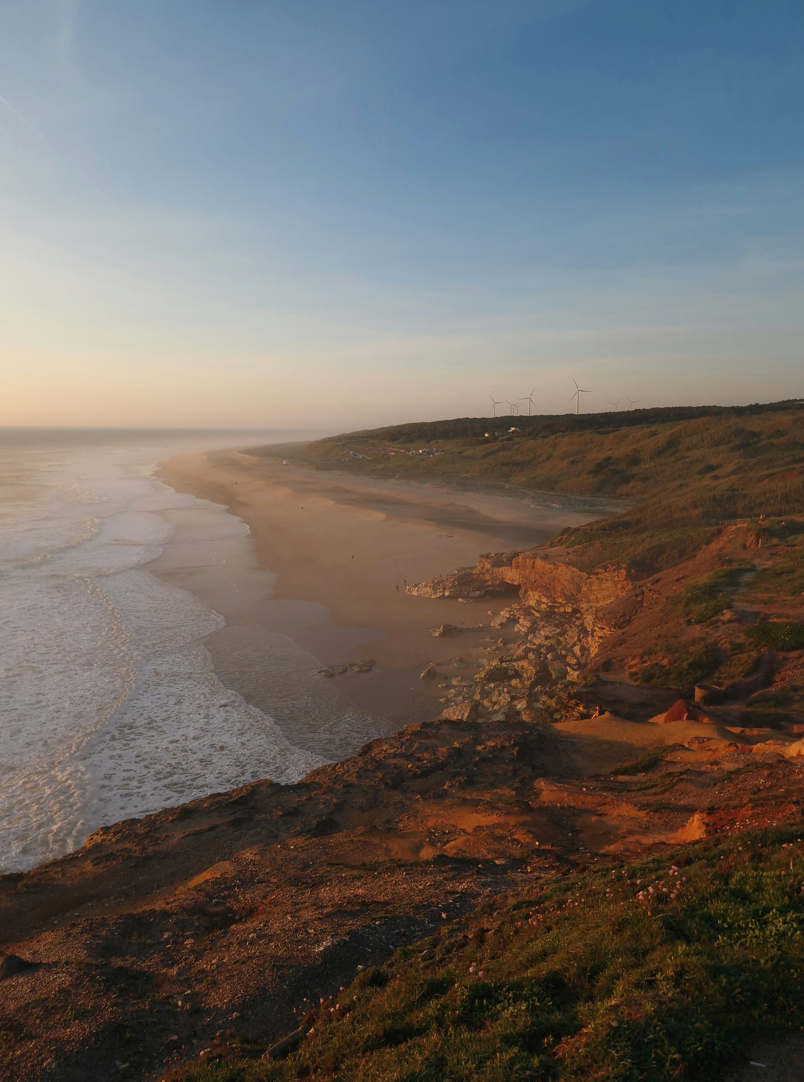 Golden sunlight casts a warm glow over a serene beach, where waves gently kiss the sandy shore, framed by rugged cliffs and lush greenery.