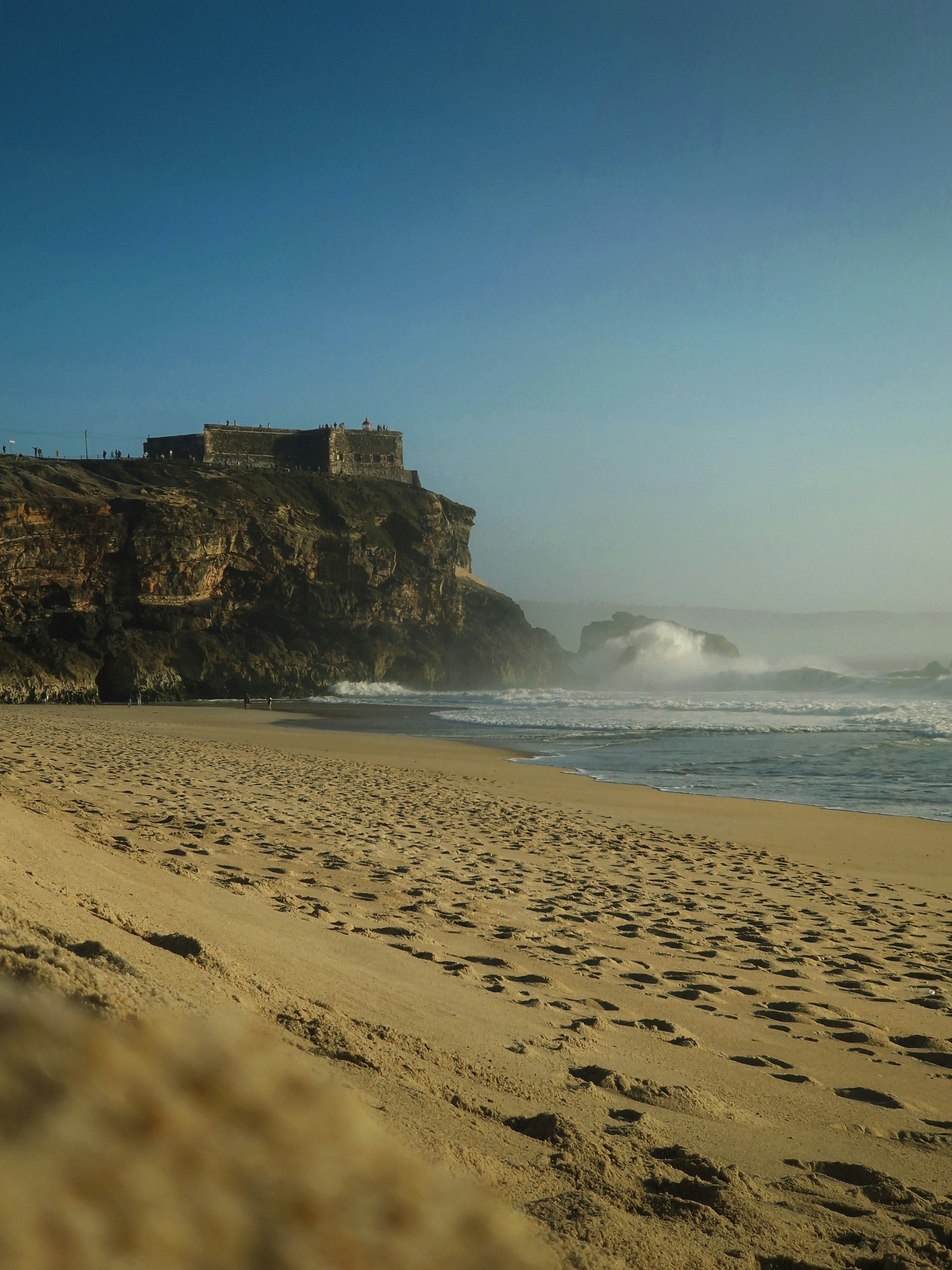 Historic fortress perched on a cliff overlooking the ocean, with gentle waves lapping at the sandy beach below.