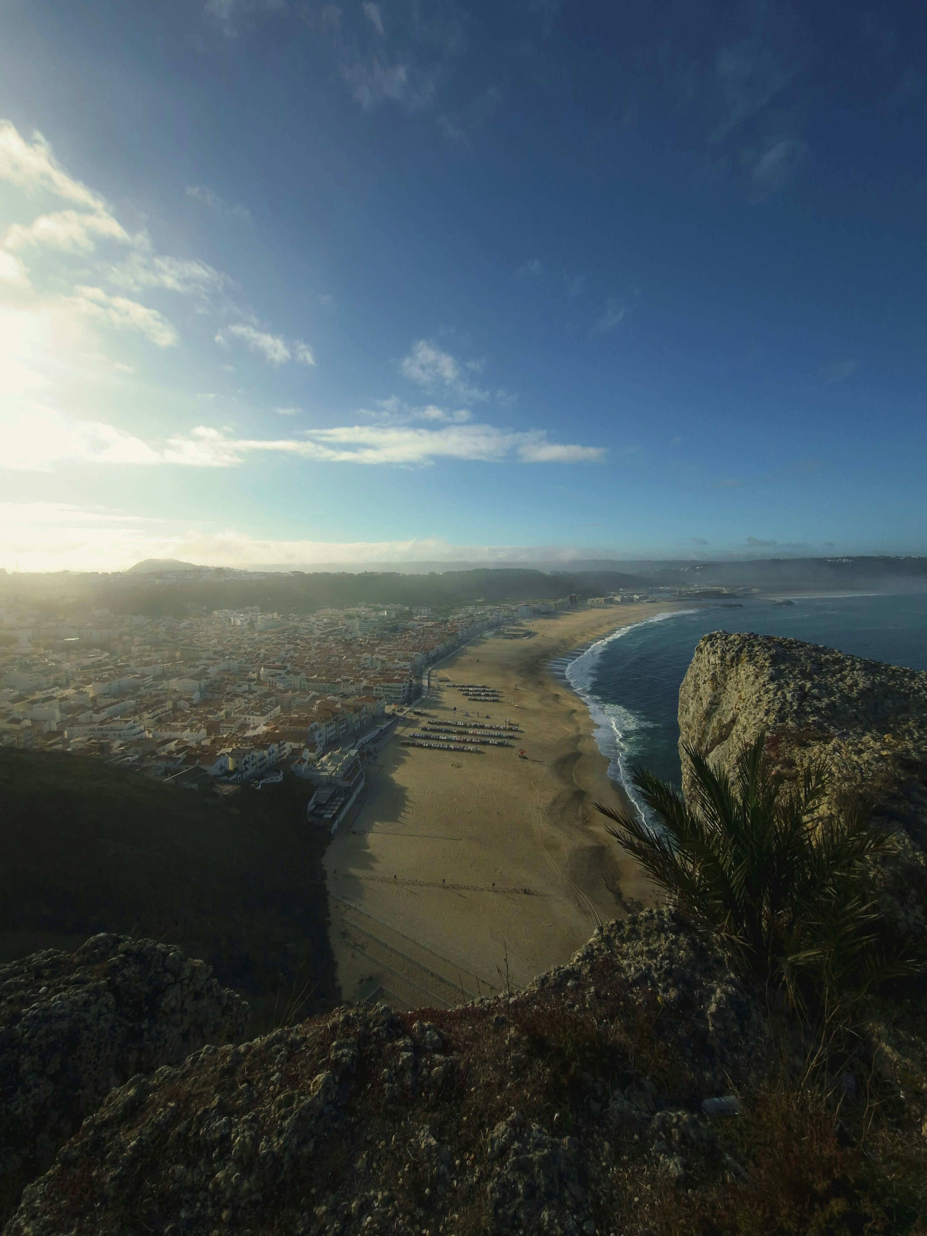 Aerial view of a sandy beach meeting the ocean, framed by rocky cliffs and a distant town. The scene captures the tranquil blend of land and sea.