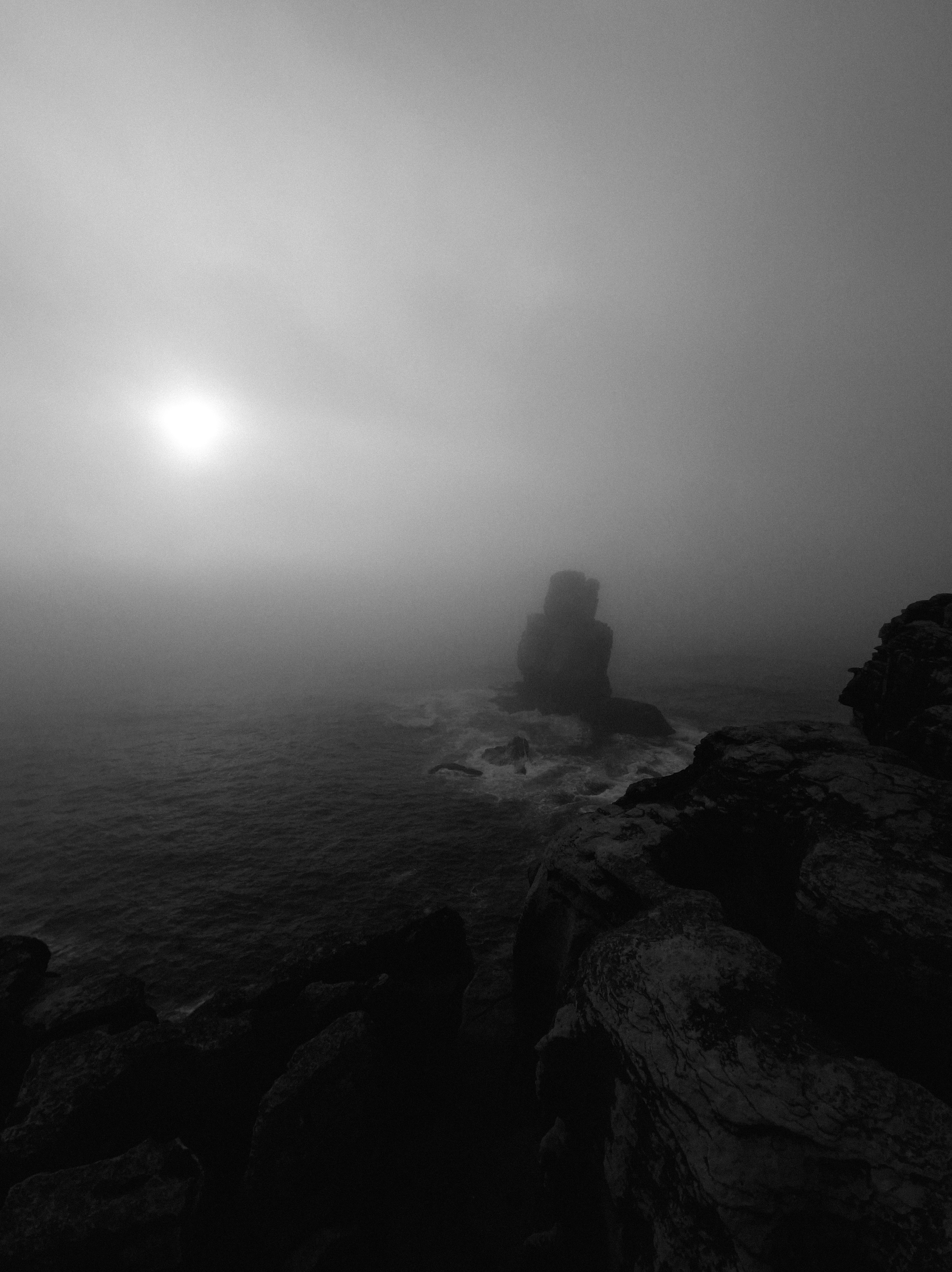 A solitary rock formation emerges from the fog, with the sun barely visible in the hazy sky. The scene evokes a sense of mystery and tranquility.