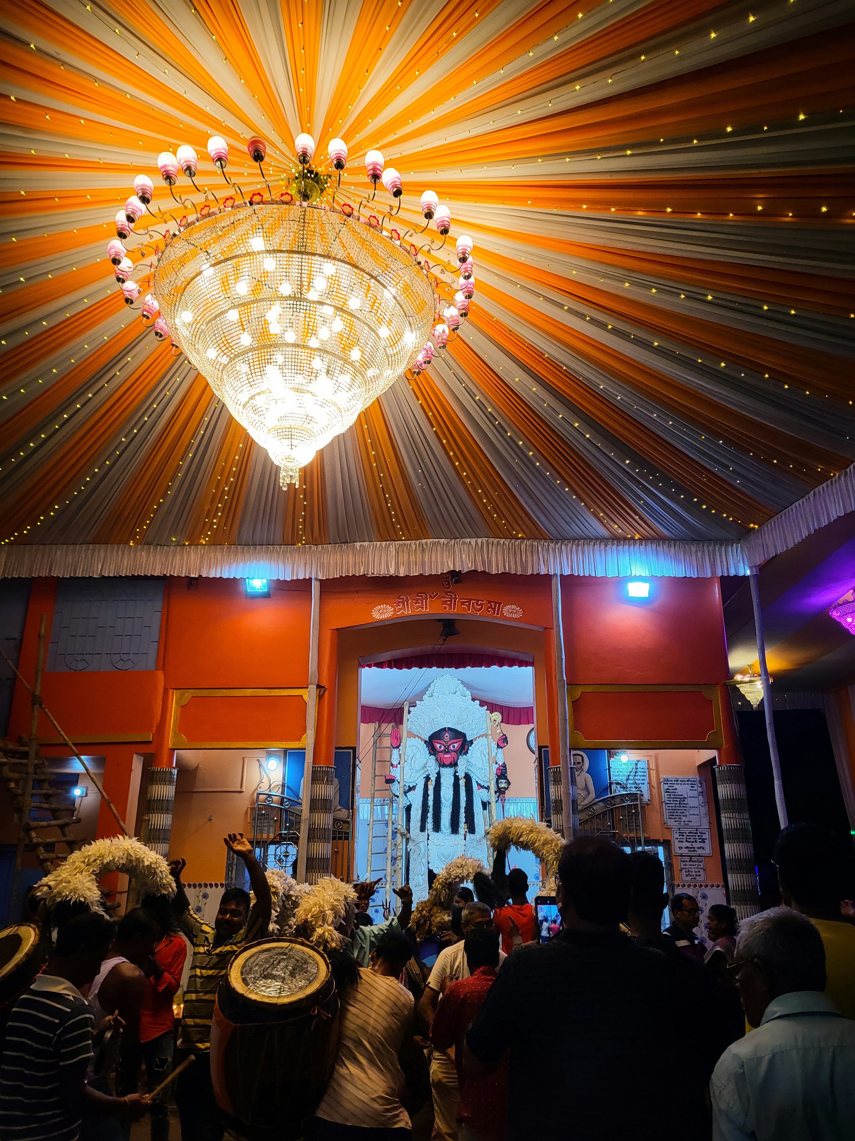 A vibrant festival scene featuring a large idol at the center, surrounded by attendees in festive attire, with a striking chandelier overhead. The colorful drapery adds to the celebratory atmosphere.