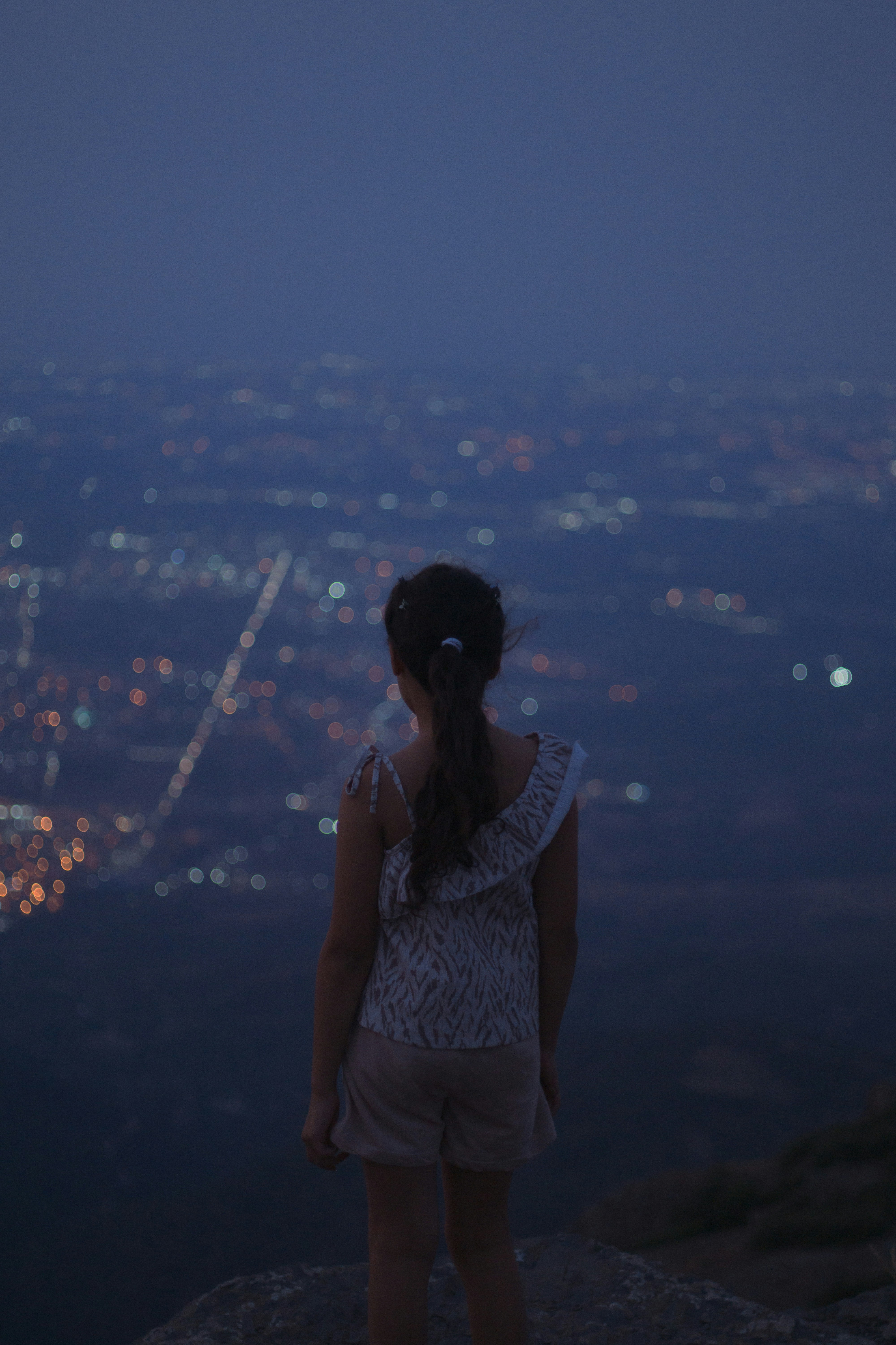 Young girl gazing at a sprawling cityscape illuminated by distant lights during twilight.