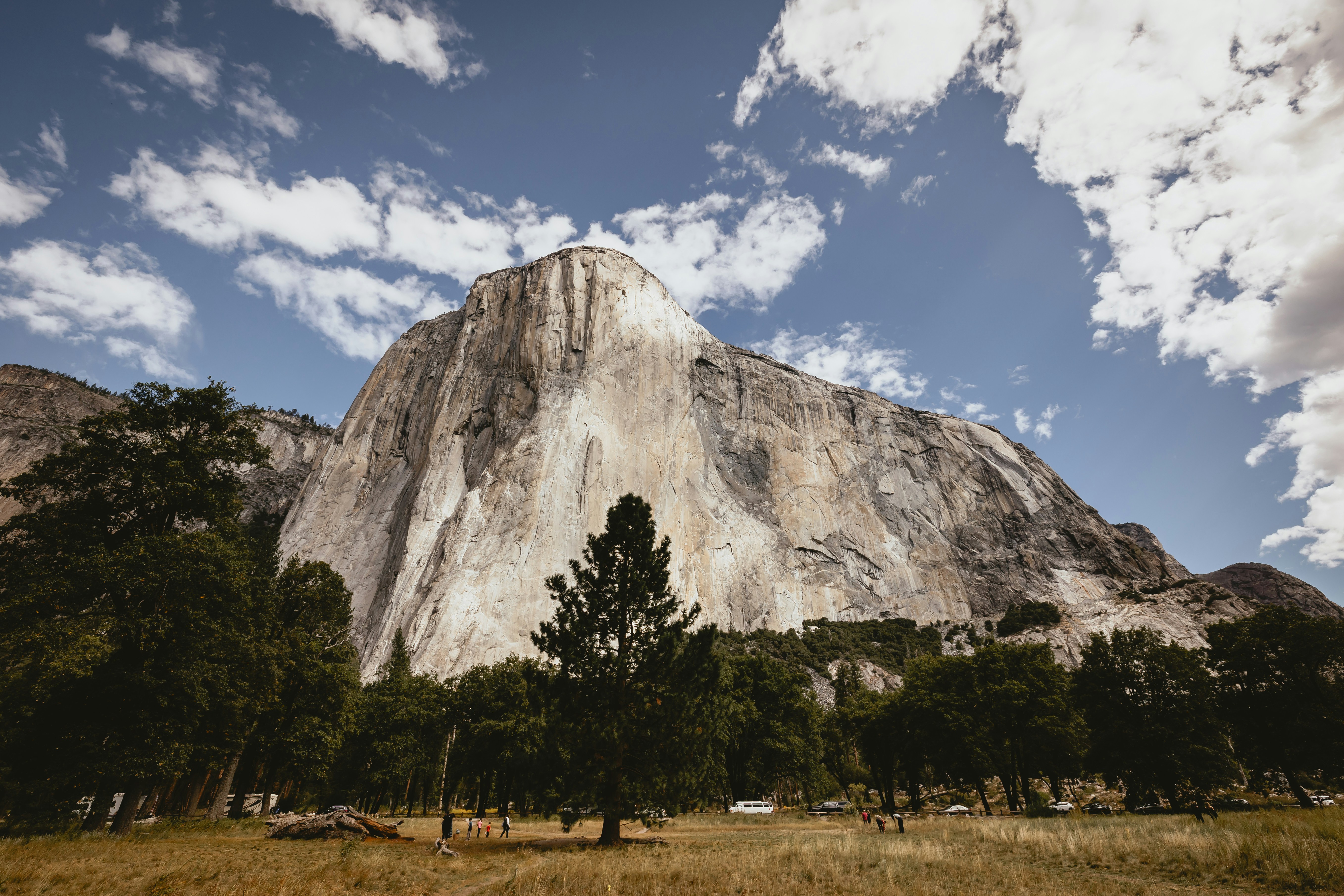 Towering granite cliff rises dramatically against a backdrop of scattered clouds and lush greenery. The scene captures the grandeur of the natural landscape.