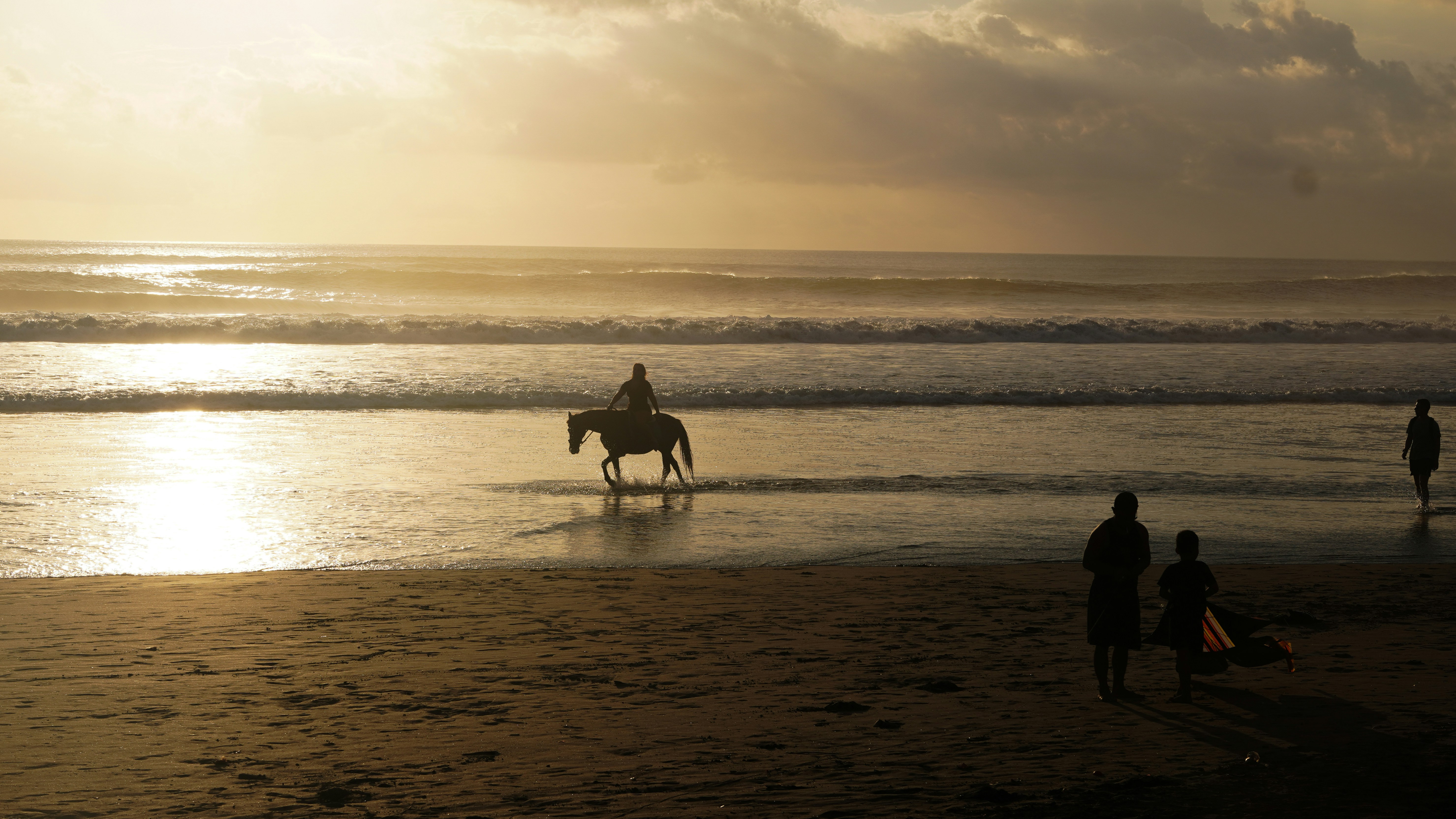 Silhouette of a rider on horseback wading through shallow waves as the sun sets, with beachgoers in the foreground.