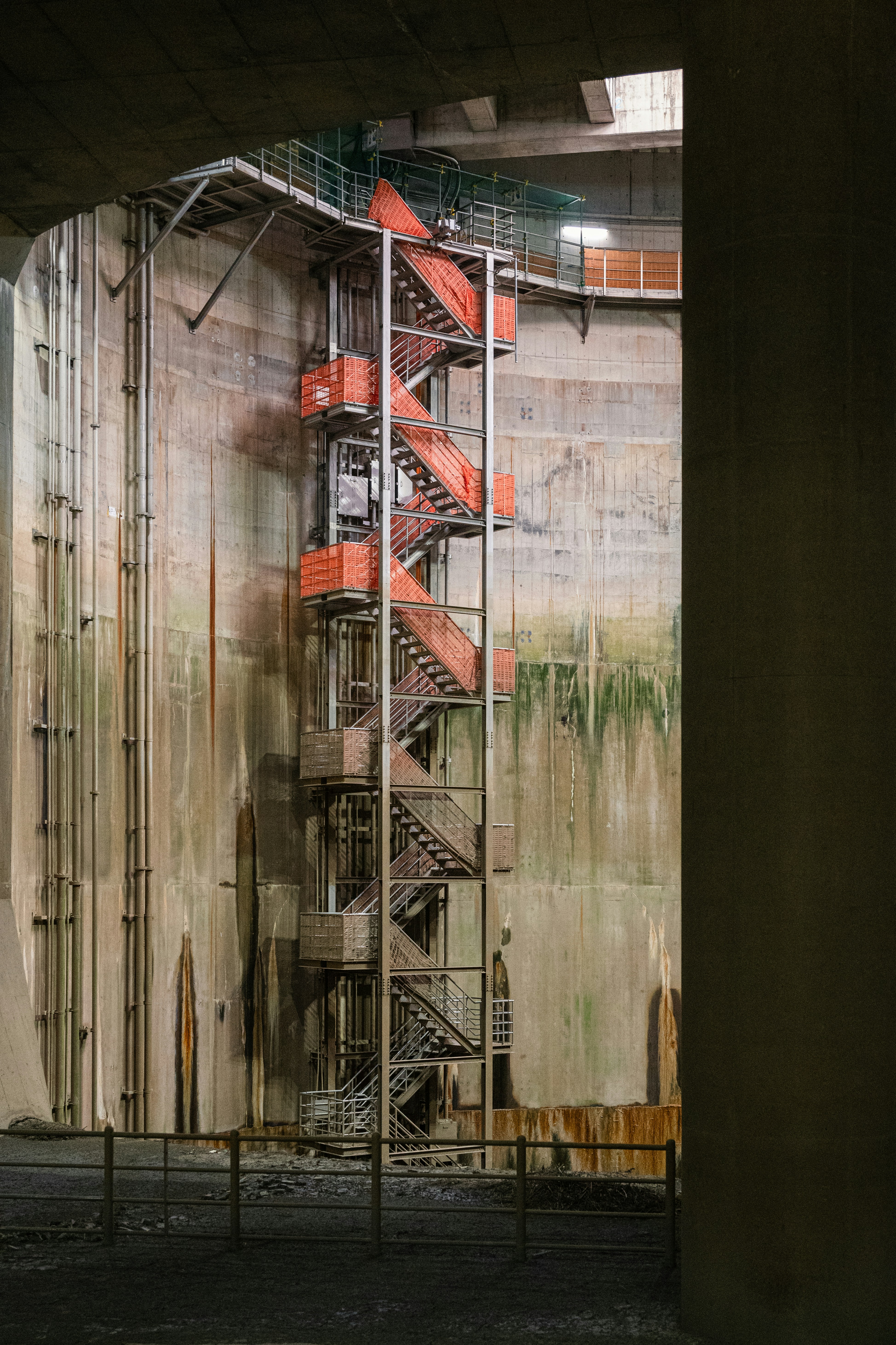A spiral staircase with orange accents rises against a weathered concrete backdrop, showcasing the interplay of industrial design and natural deterioration.