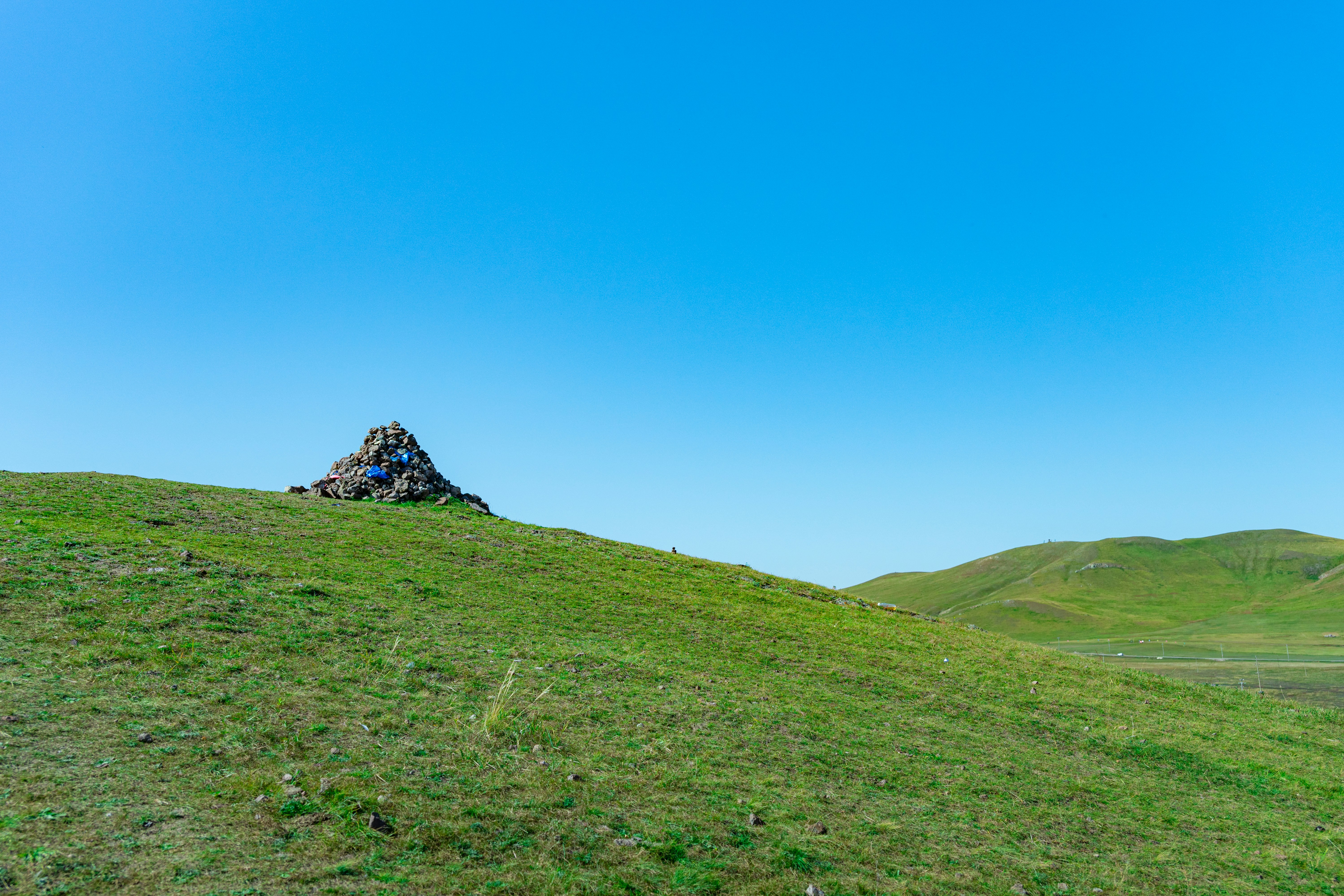 A small stone mound rises on a grassy hill under a clear blue sky, hinting at historical significance in a serene landscape.
