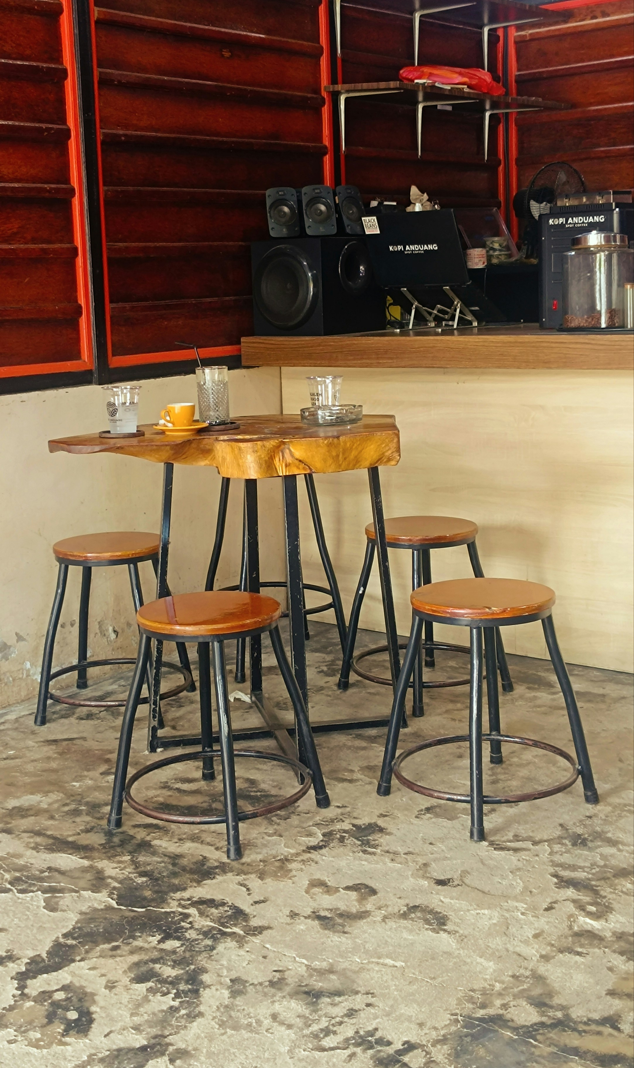 A wooden table with metal legs surrounded by stools in a quaint café setting, featuring a simple bar counter and warm wooden accents.