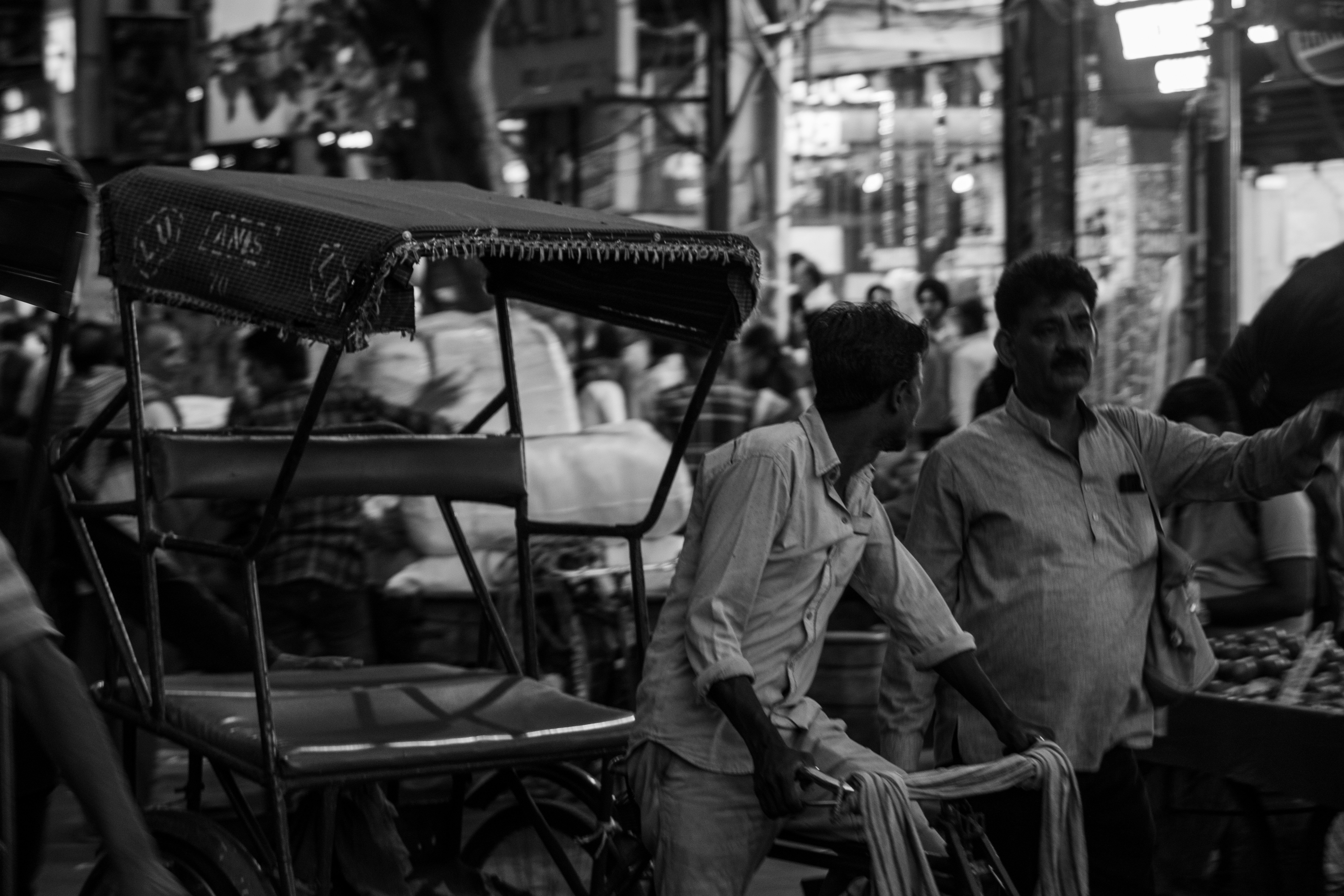 Hello from the streets of Chandni Chowk, one of Delhi’s oldest and busiest marketplaces, filled with hand-pulled rickshaws and streams of people moving through narrow lanes. This photo is a part of a series of Black and White photos that I took to try to mimic the time of black and white film. I hope you like it and you find it useful in your next project. | Two men stand near a rickshaw in a busy street.