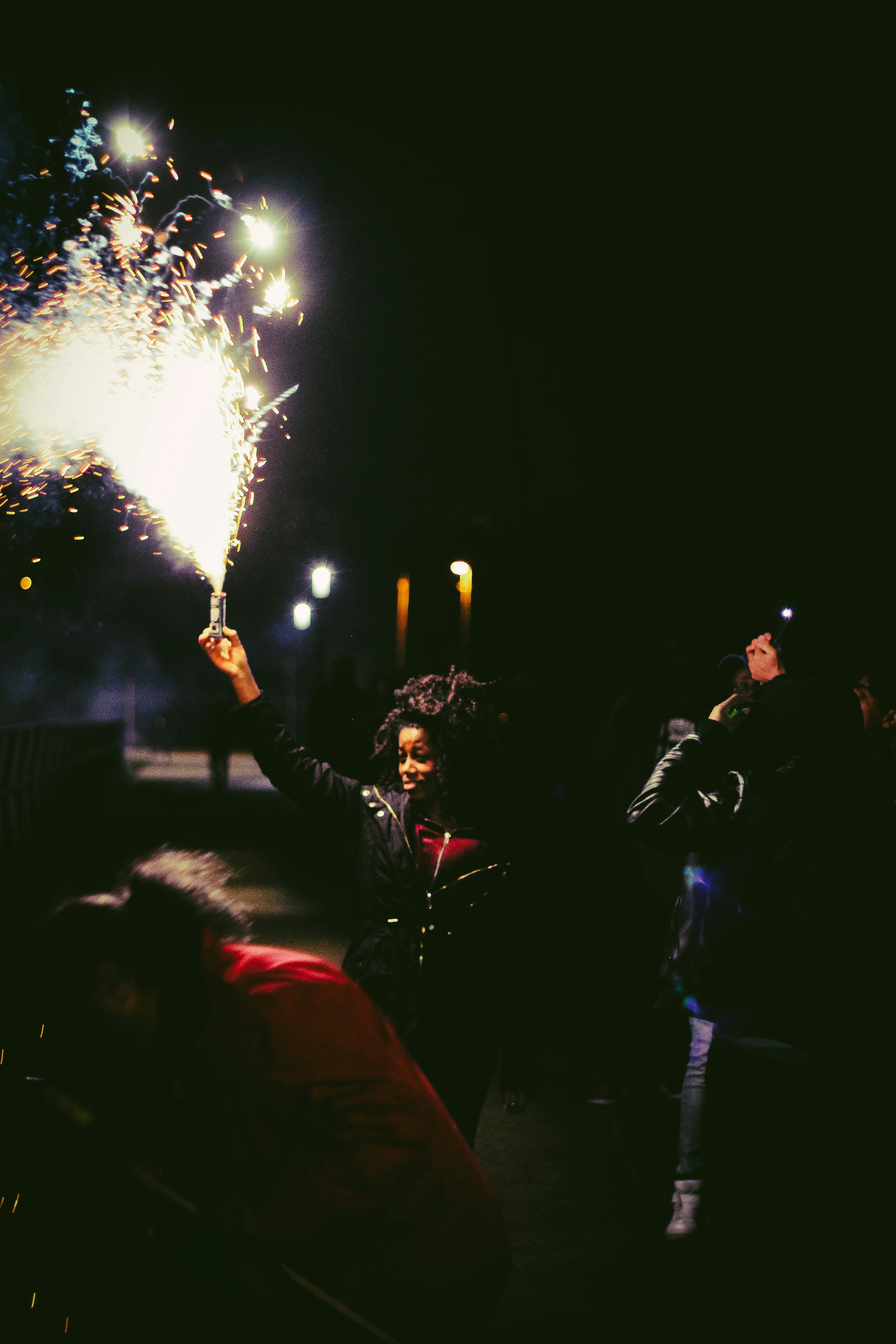 Person holding a sparkler, surrounded by friends in a festive nighttime atmosphere. The scene captures the joy and excitement of celebration.