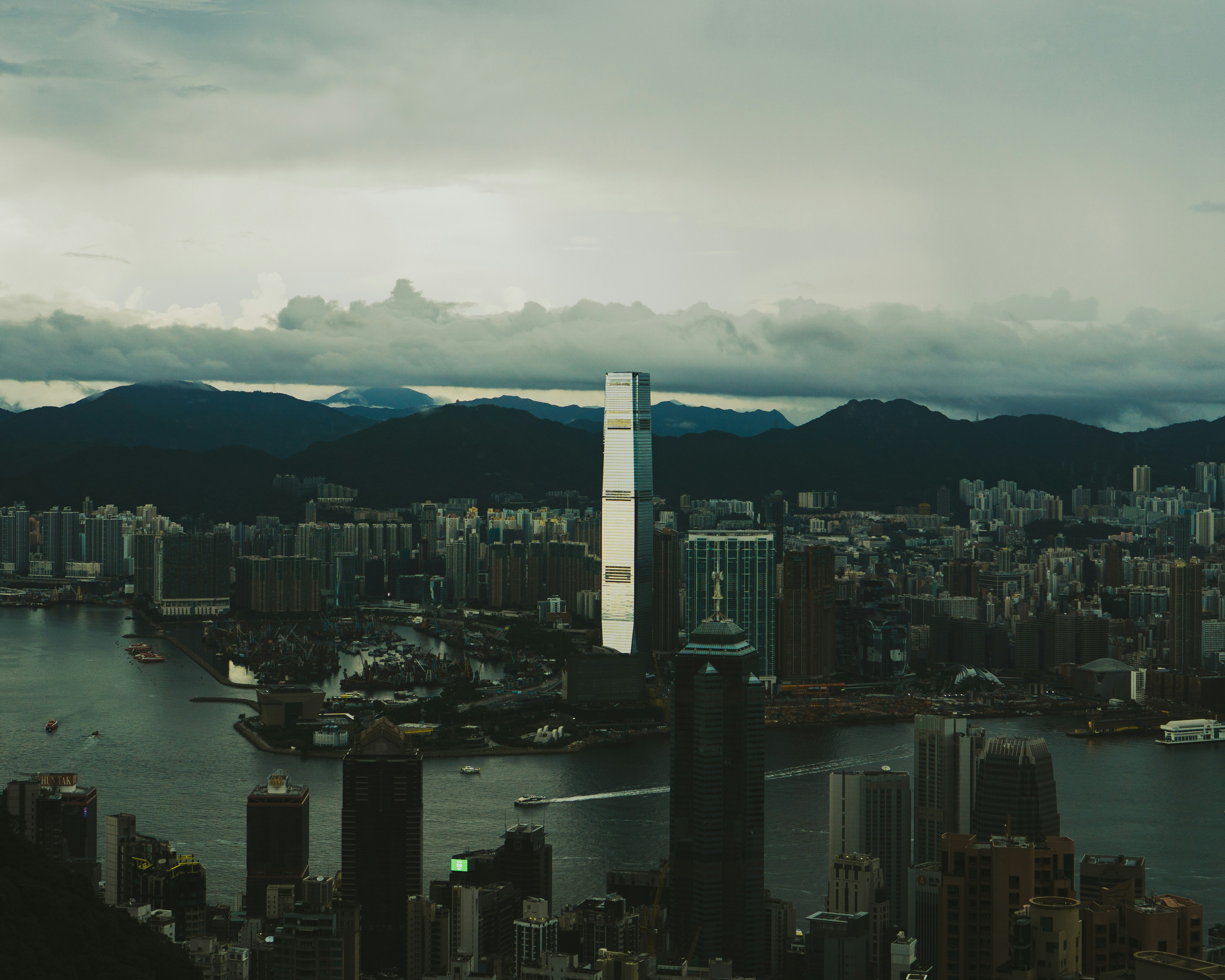 The International Commerce Centre (ICC), Hong Kong's tallest skyscraper, pierces a dramatic, overcast sky, its gleaming facade catching the light. Viewed from a high vantage point, the landmark tower dominates the West Kowloon landscape, standing in stark contrast to the dark, moody waters of Victoria Harbour and the silhouetted mountains behind it. | Tall modern skyscraper in a dense city skyline