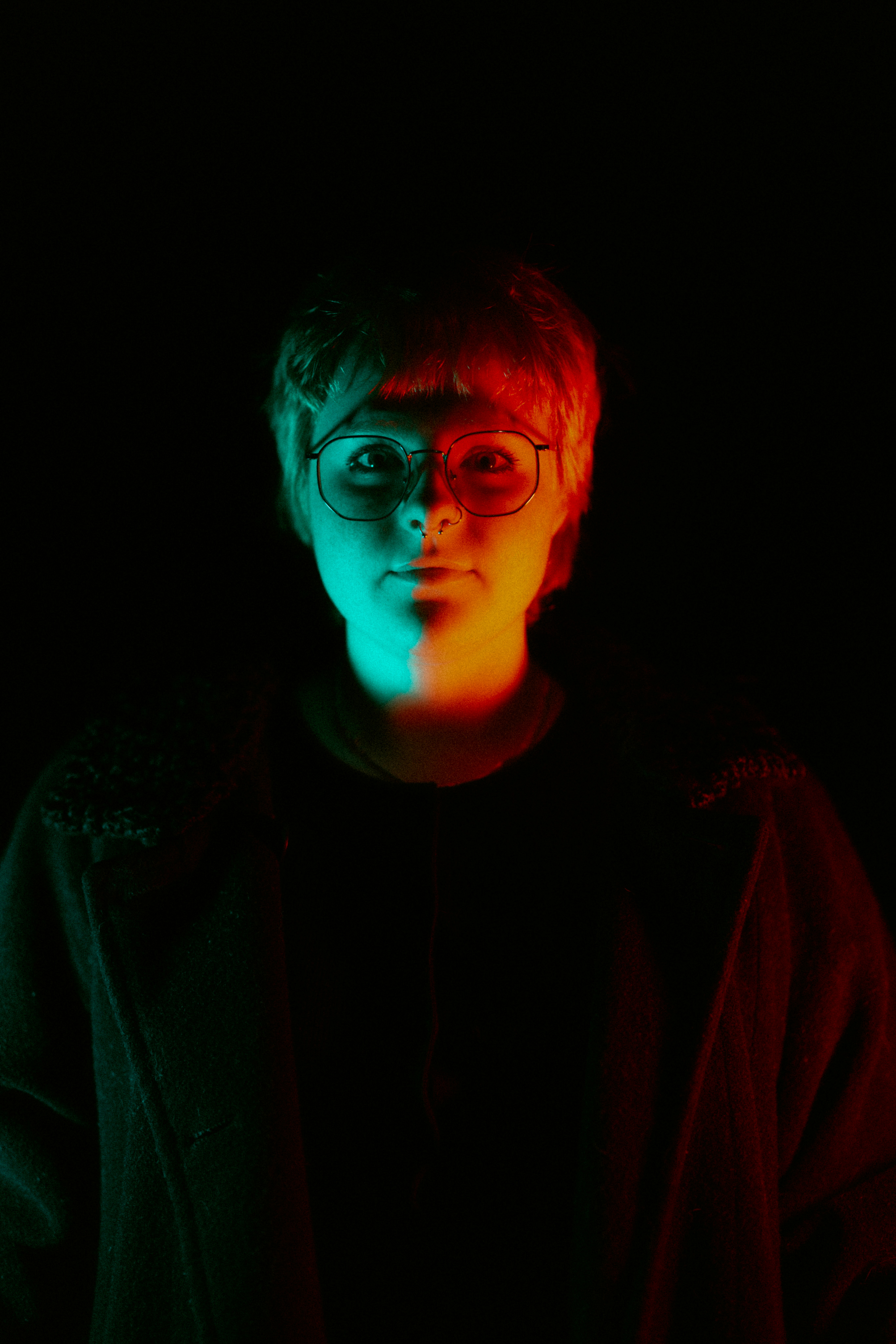 A figure stands against a black background, illuminated by dramatic teal and red lighting, creating a striking visual contrast. The subject's glasses and expression add depth to the scene.