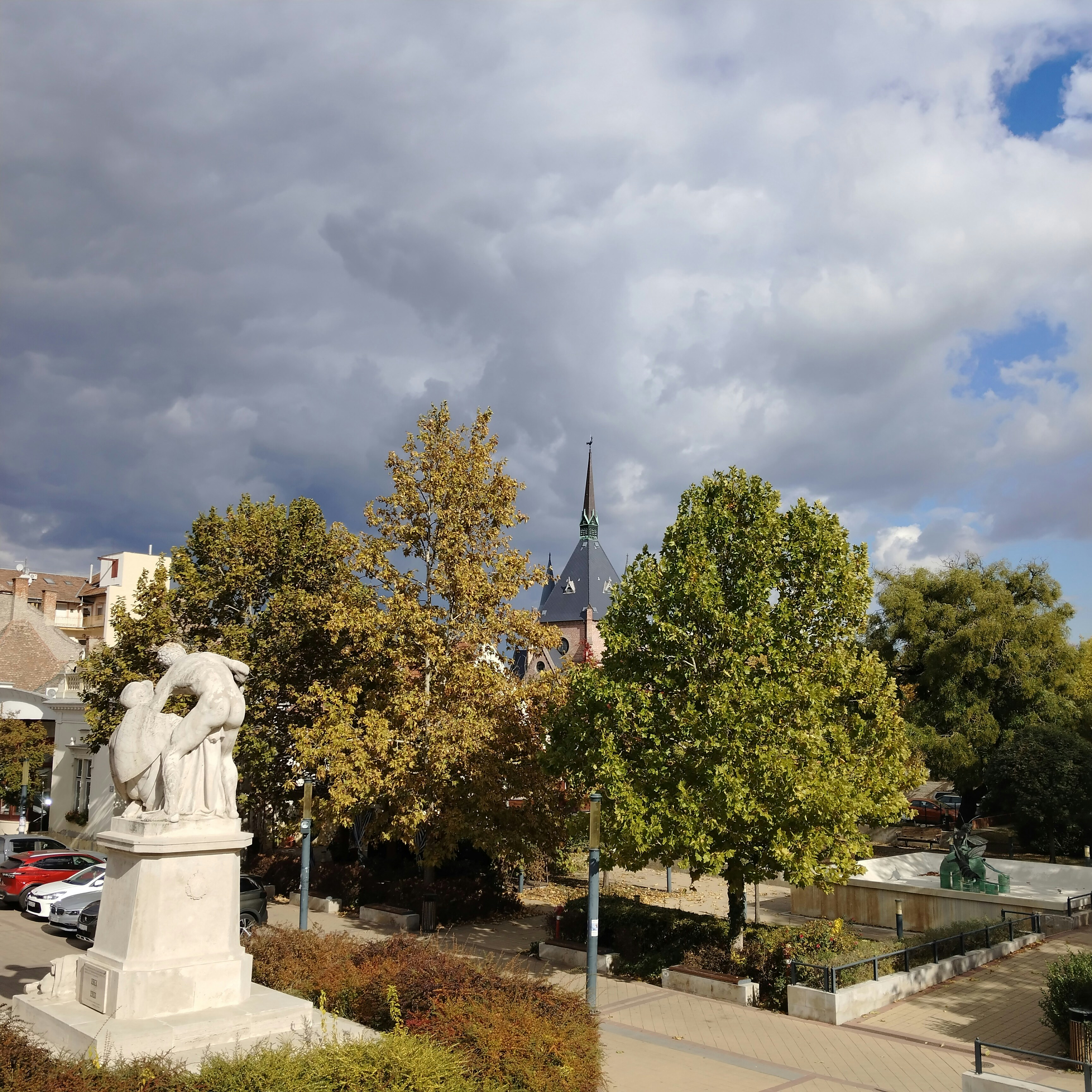 A serene statue surrounded by vibrant autumn foliage, capturing a moment of tranquility in a public square. The backdrop features a mix of historical architecture and dramatic clouds.