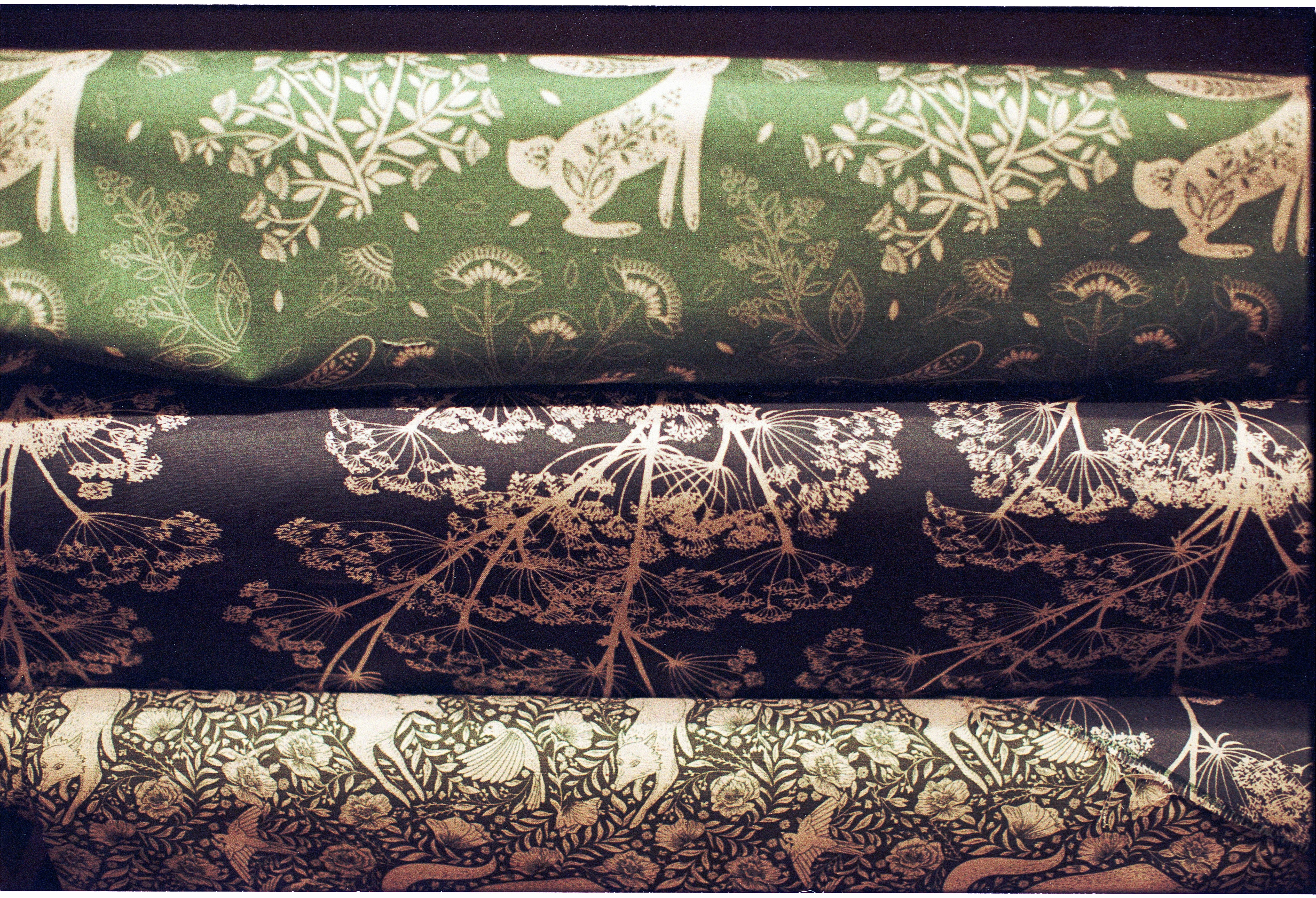 Three rolls of patterned fabric showcasing intricate botanical designs with animal motifs in varying color schemes. The textures and details create a visually appealing display.