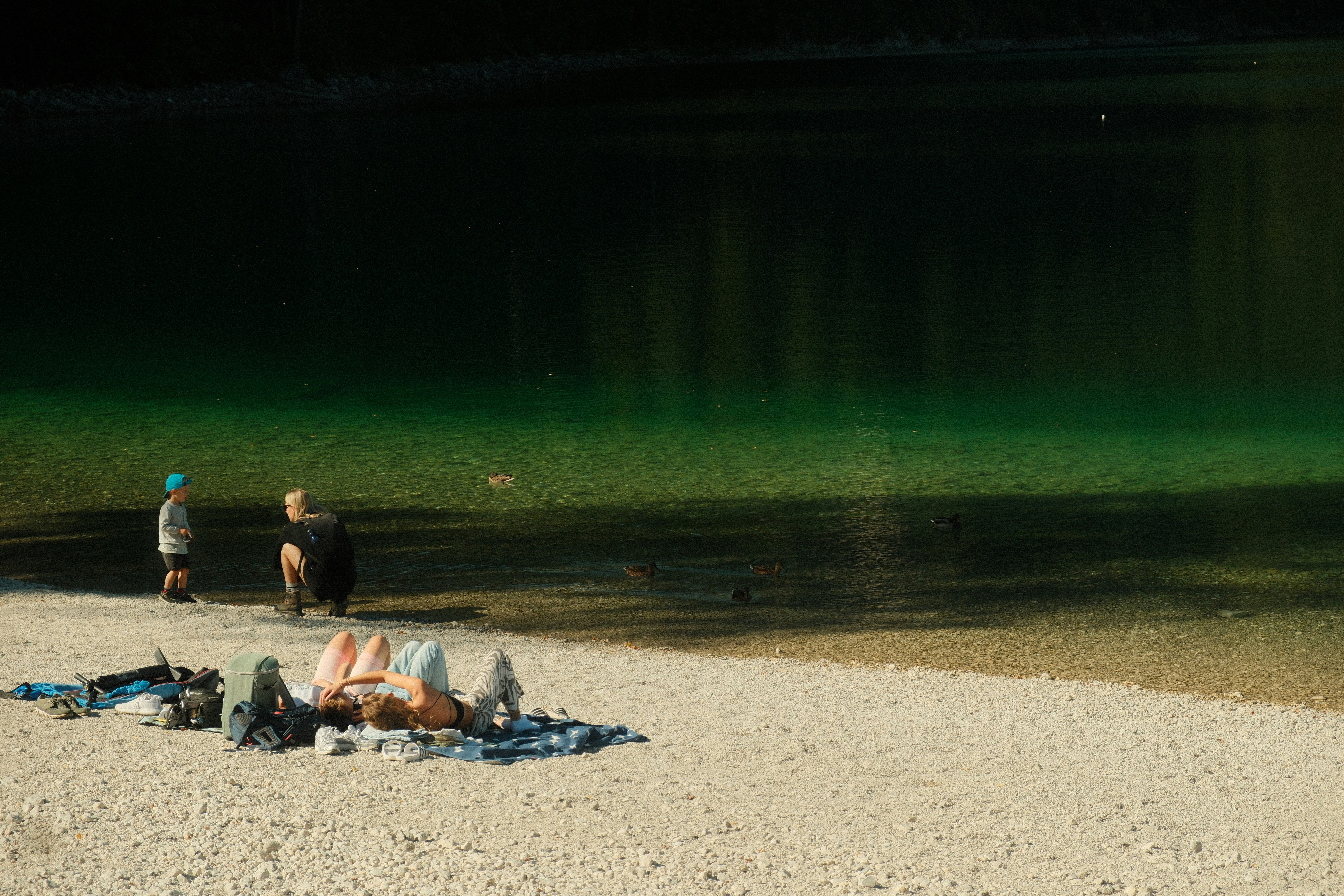 A tranquil lakeside scene featuring a child exploring the shoreline while an adult relaxes nearby. The crystal-clear water reflects the surrounding nature.