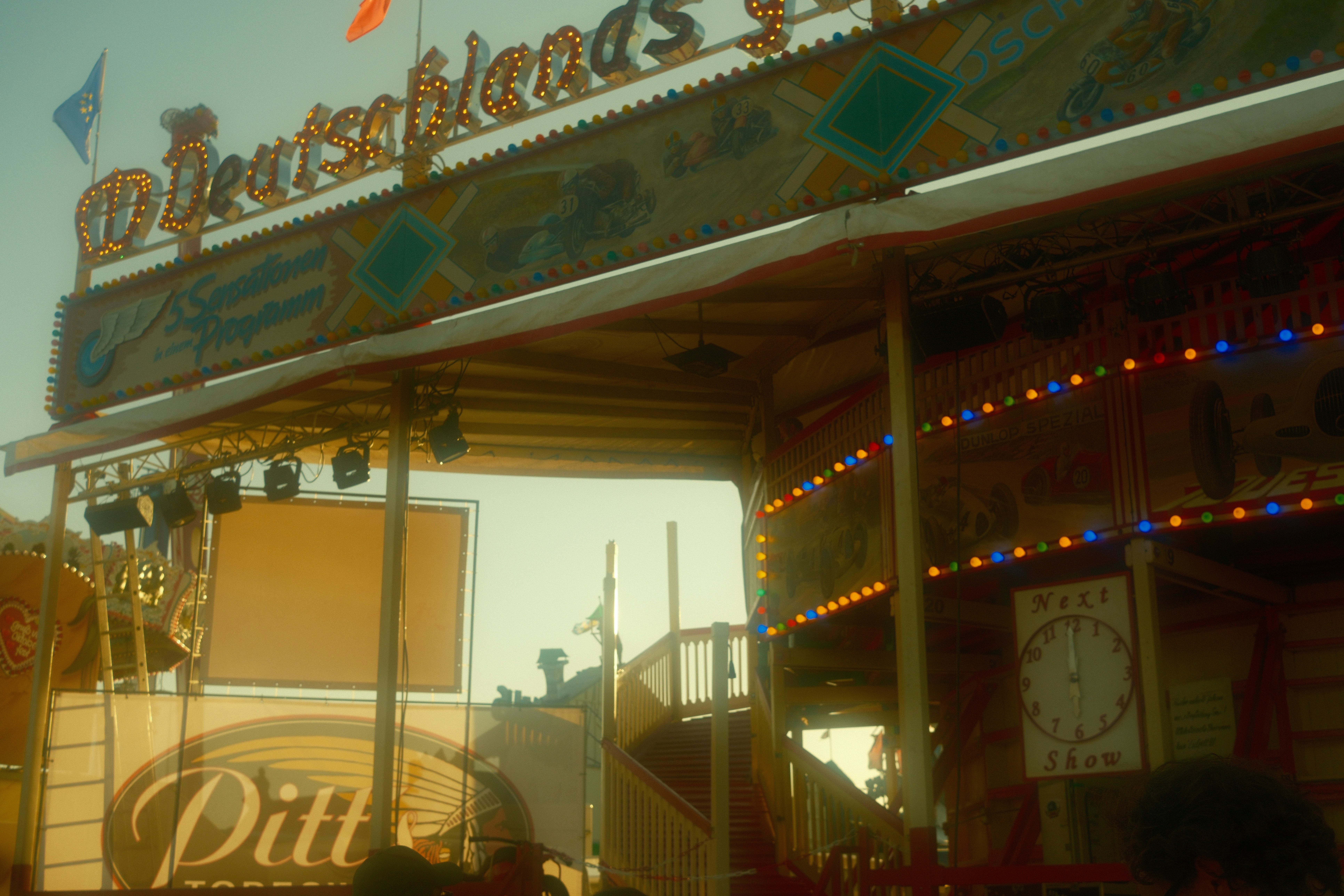 Pastel dusk over a retro carnival facade: marquee bulbs, a stairway entrance, and a vintage clock.