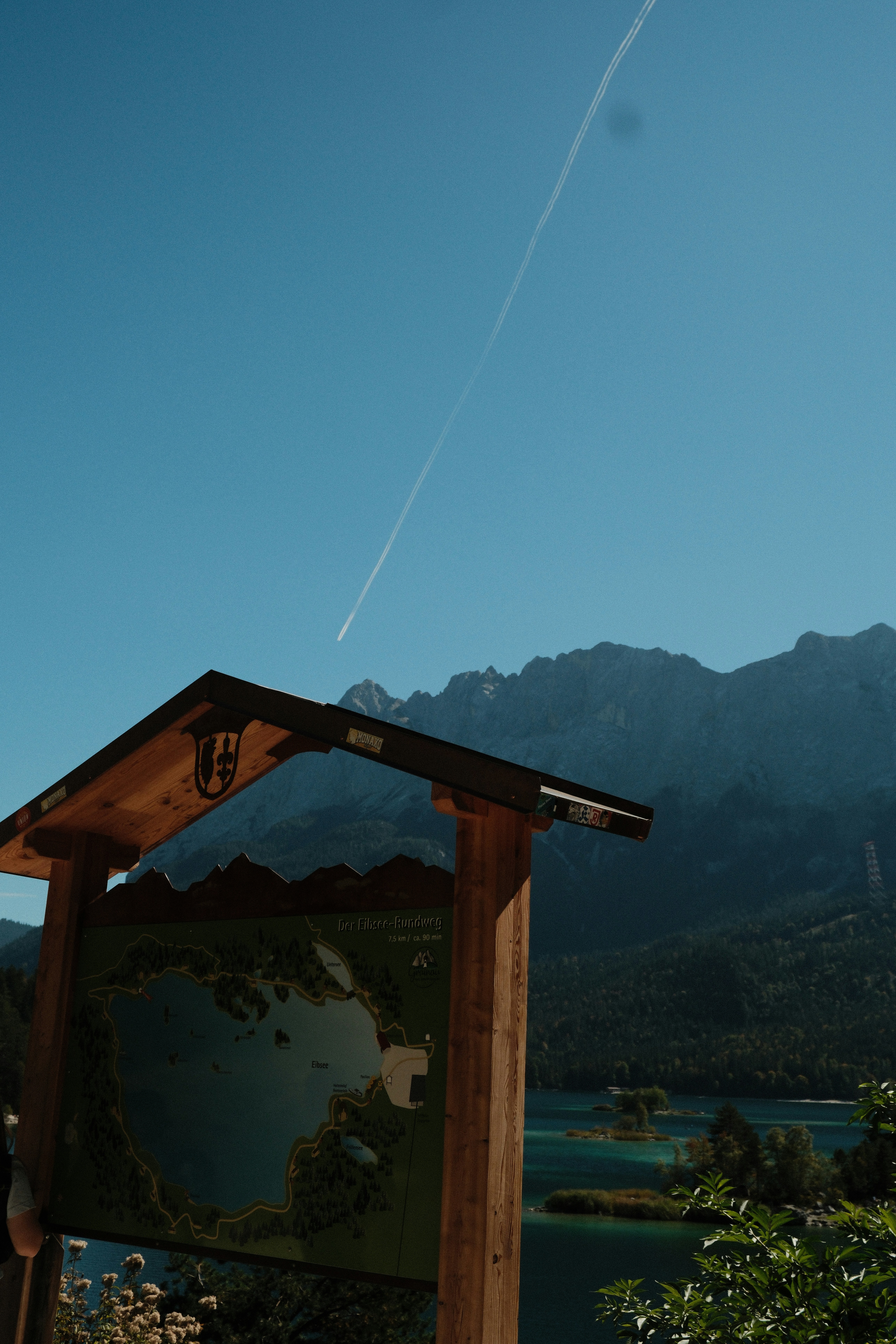 A wooden informational sign stands in the foreground, detailing the surrounding lake and mountains under a clear blue sky with a jet trail above.