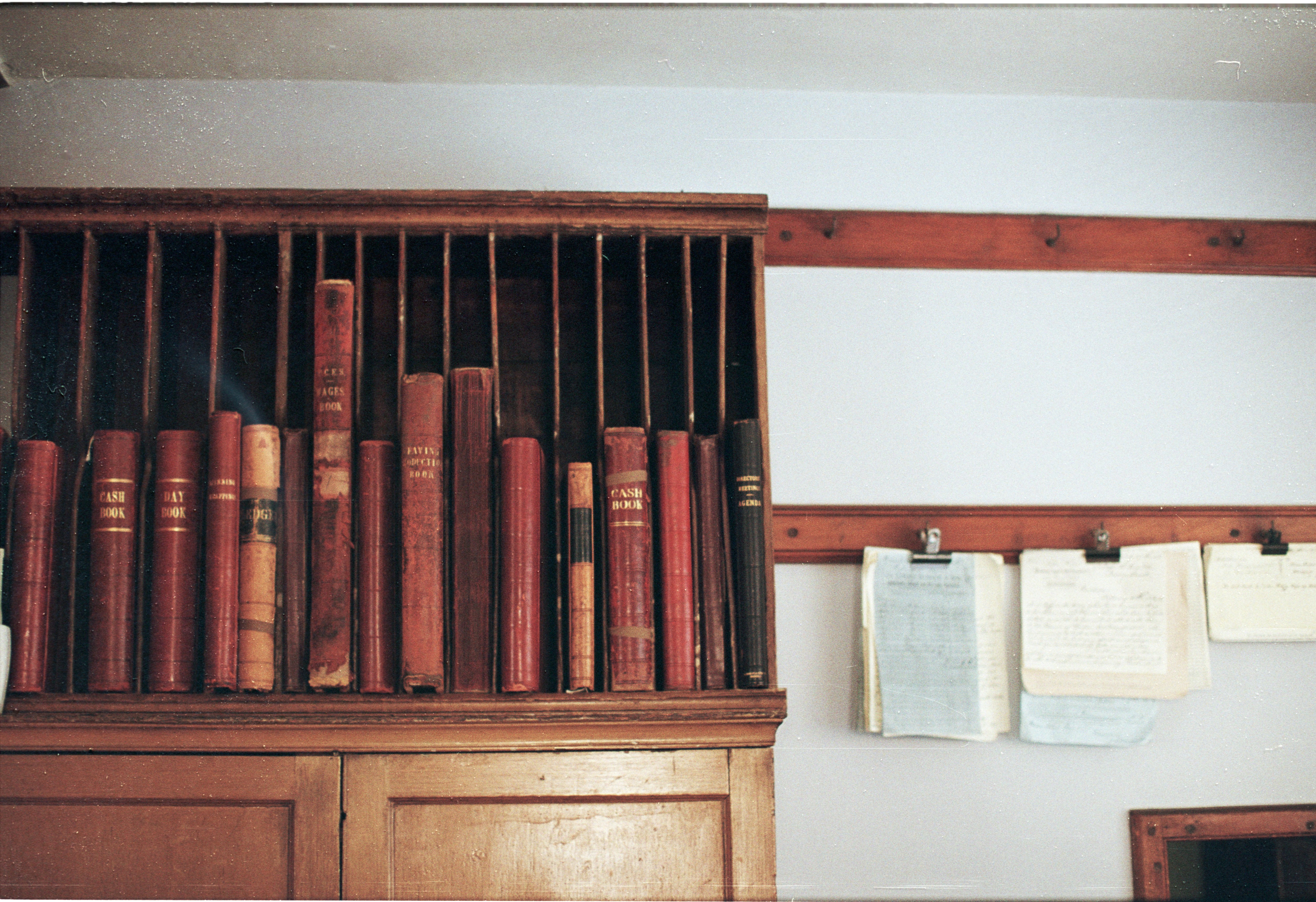 A collection of antique books arranged in a wooden shelf, complemented by hanging documents on the wall. The scene evokes a sense of nostalgia and scholarly pursuit.