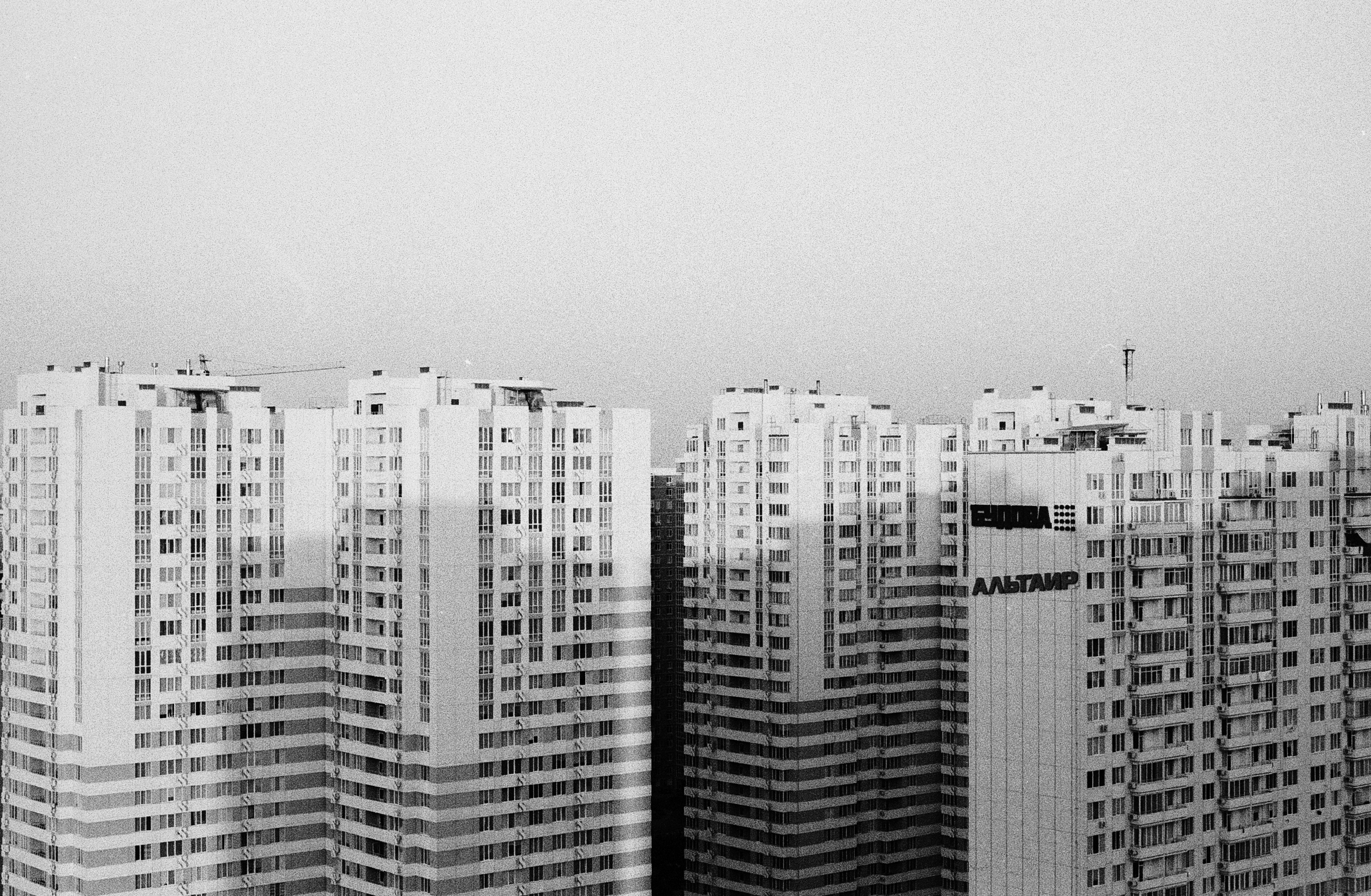 A minimalist monochrome image captures the imposing, dense verticality of a cluster of modern high-rise apartment buildings. The structures dominate the frame, their repeating patterns of windows and horizontal stripes creating a strong geometric rhythm. The uniform white or light-colored facades are contrasted by the deep shadows in the window recesses and between the buildings. A sign with the word "АЛТАИР" (ALTAIR) is visible on the side of one tower. The sky above is a bright, featureless expanse, emphasizing the scale and sheer density of the urban architecture. | Modern apartment buildings against a bright sky