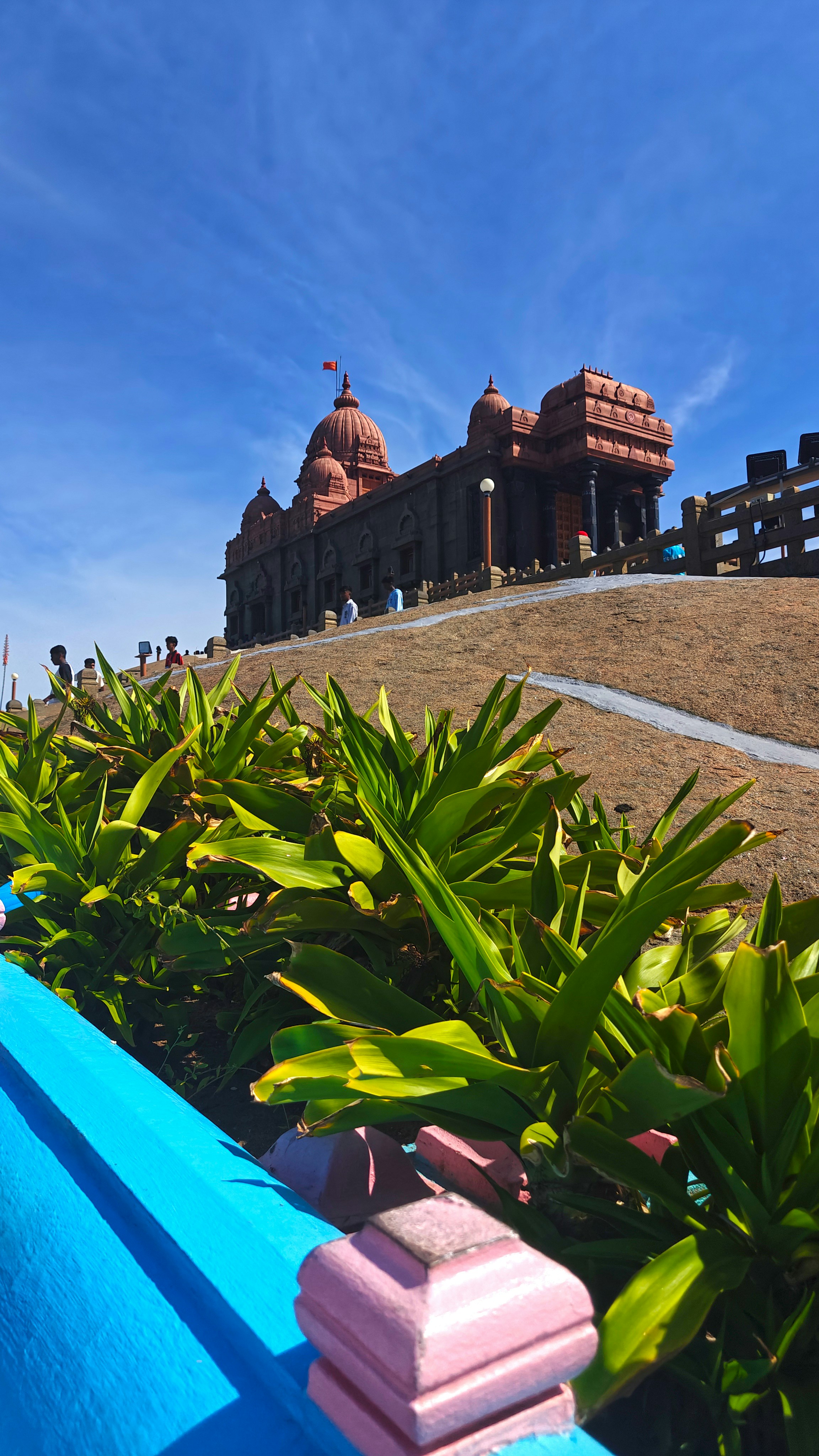 A majestic temple rises against a clear blue sky, framed by vibrant greenery and colorful railings. Visitors ascend the path, adding life to the historic scene.