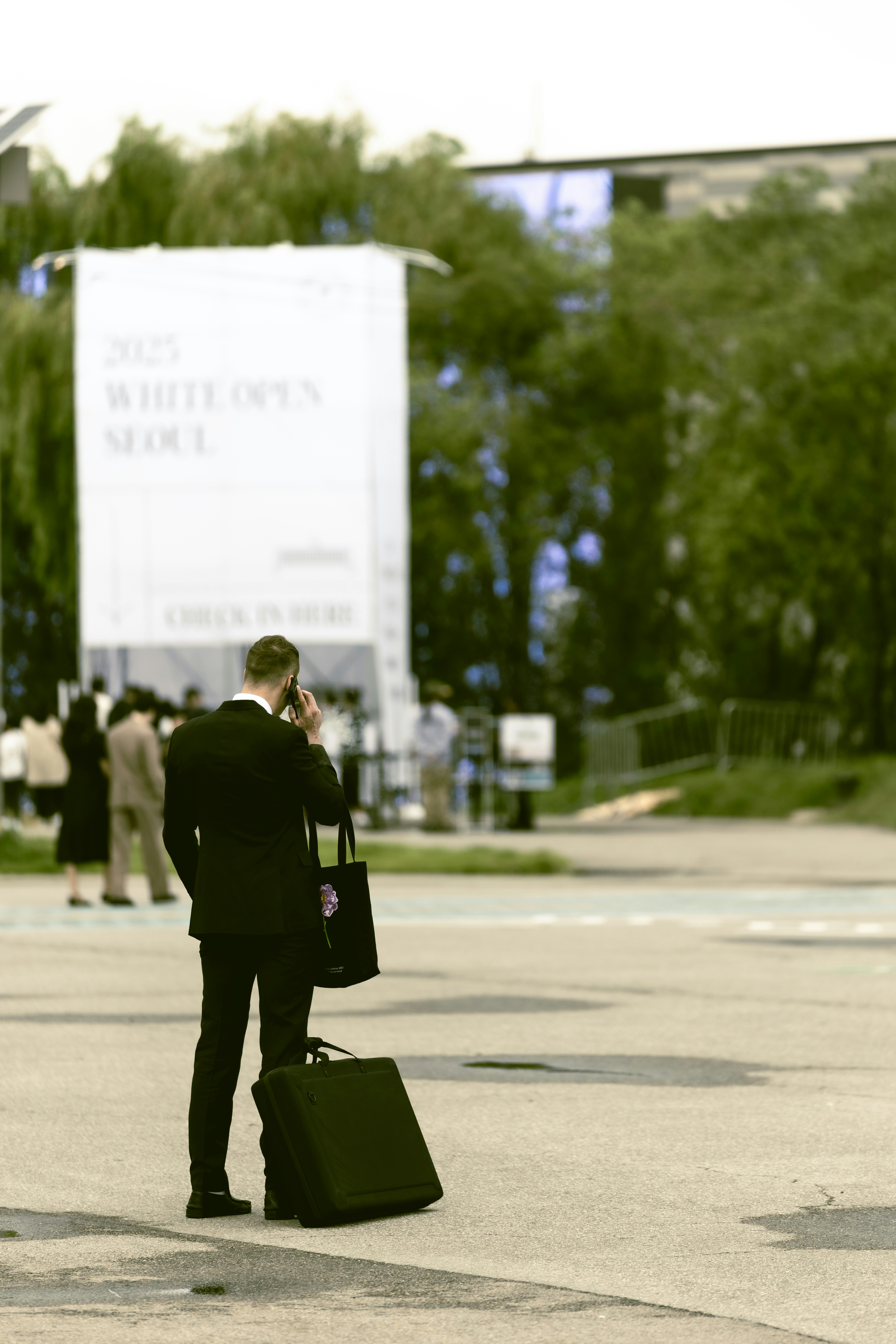 A businessman in a suit stands with a suitcase, engaged in a phone call, as a large banner for an upcoming event looms in the background.