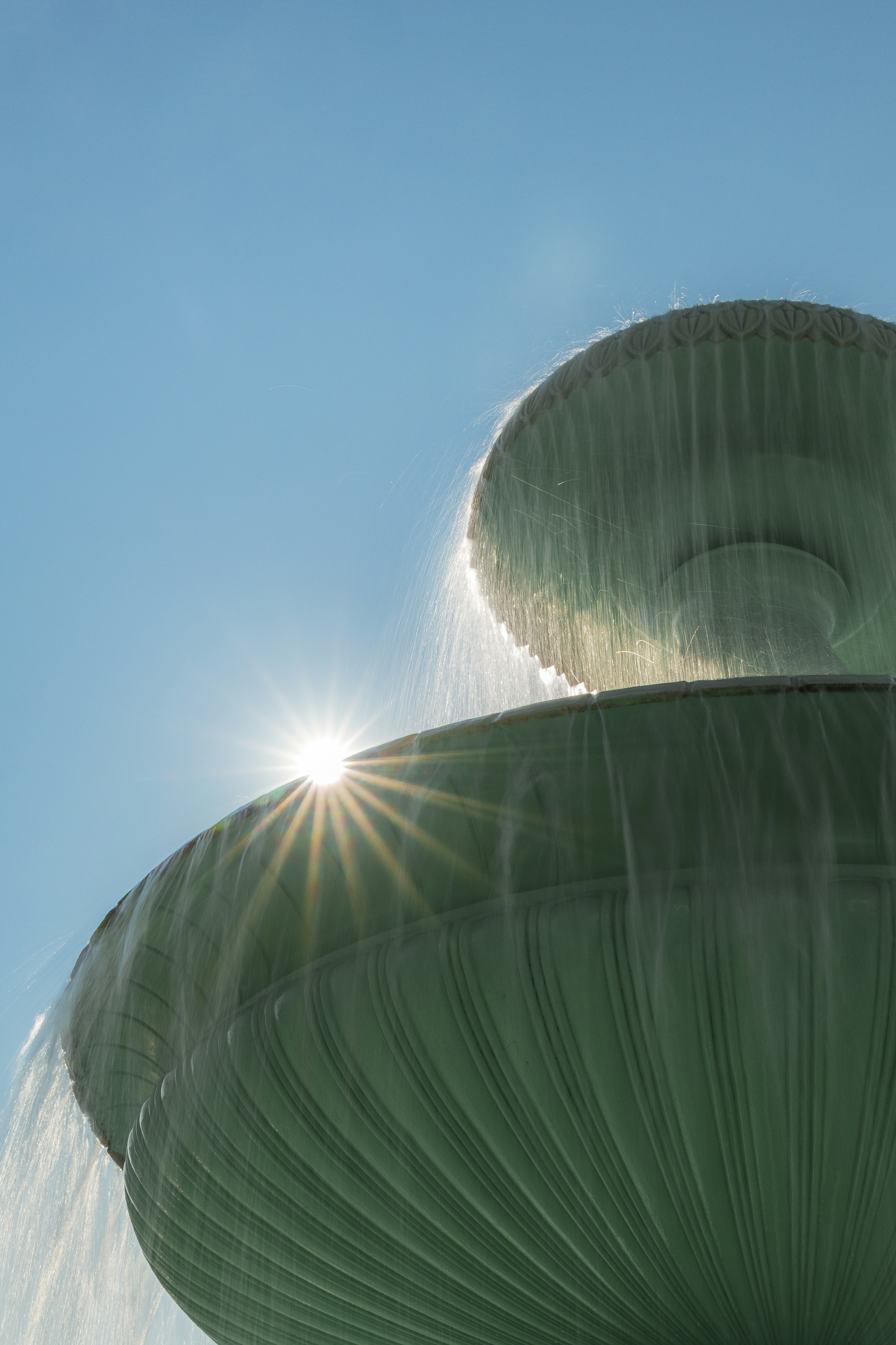 Elegant fountain with cascading water illuminated by sunlight, creating a mesmerizing display against a clear blue sky.