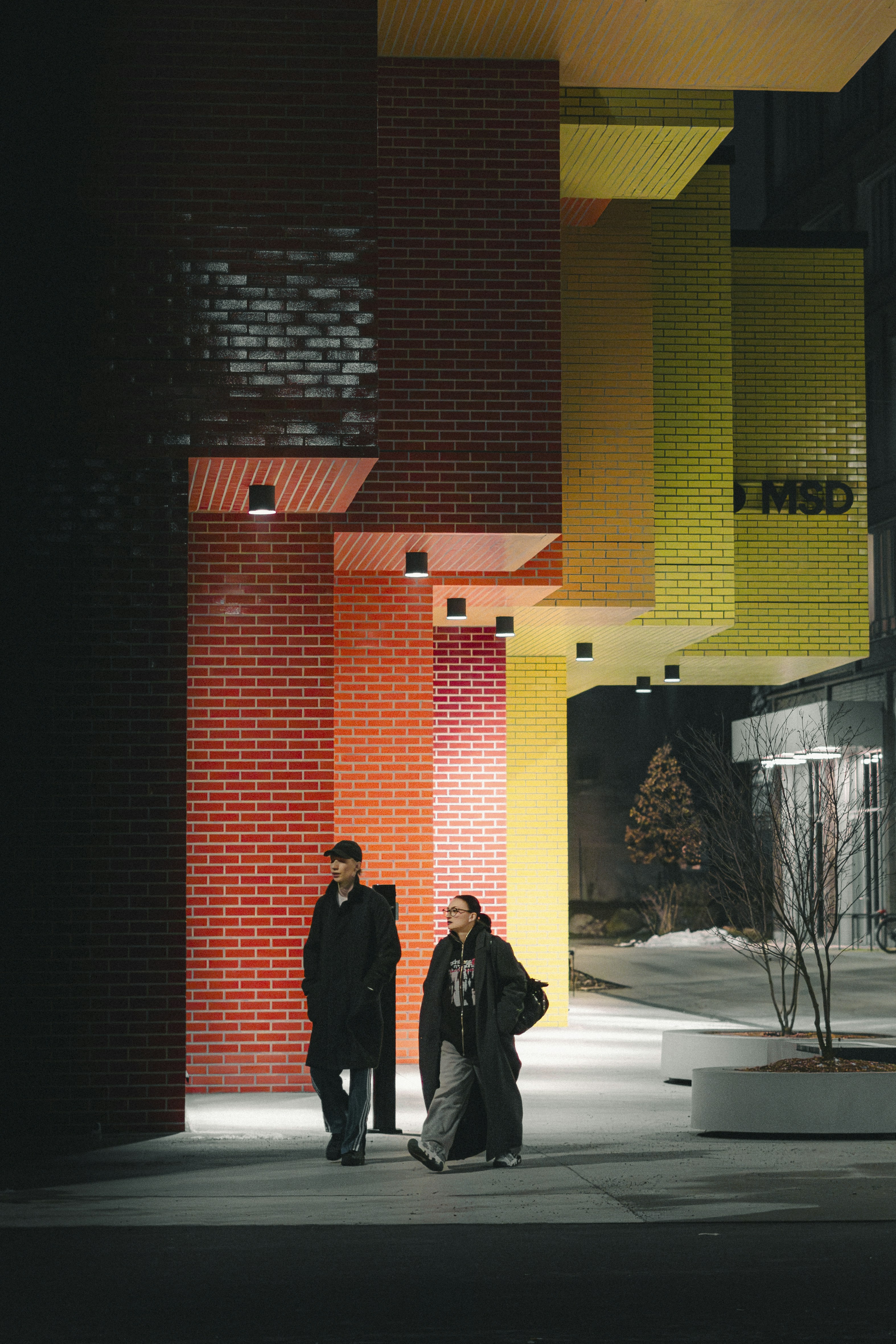 Two figures stroll beneath vibrant, illuminated architecture, showcasing a blend of modern design and urban life.