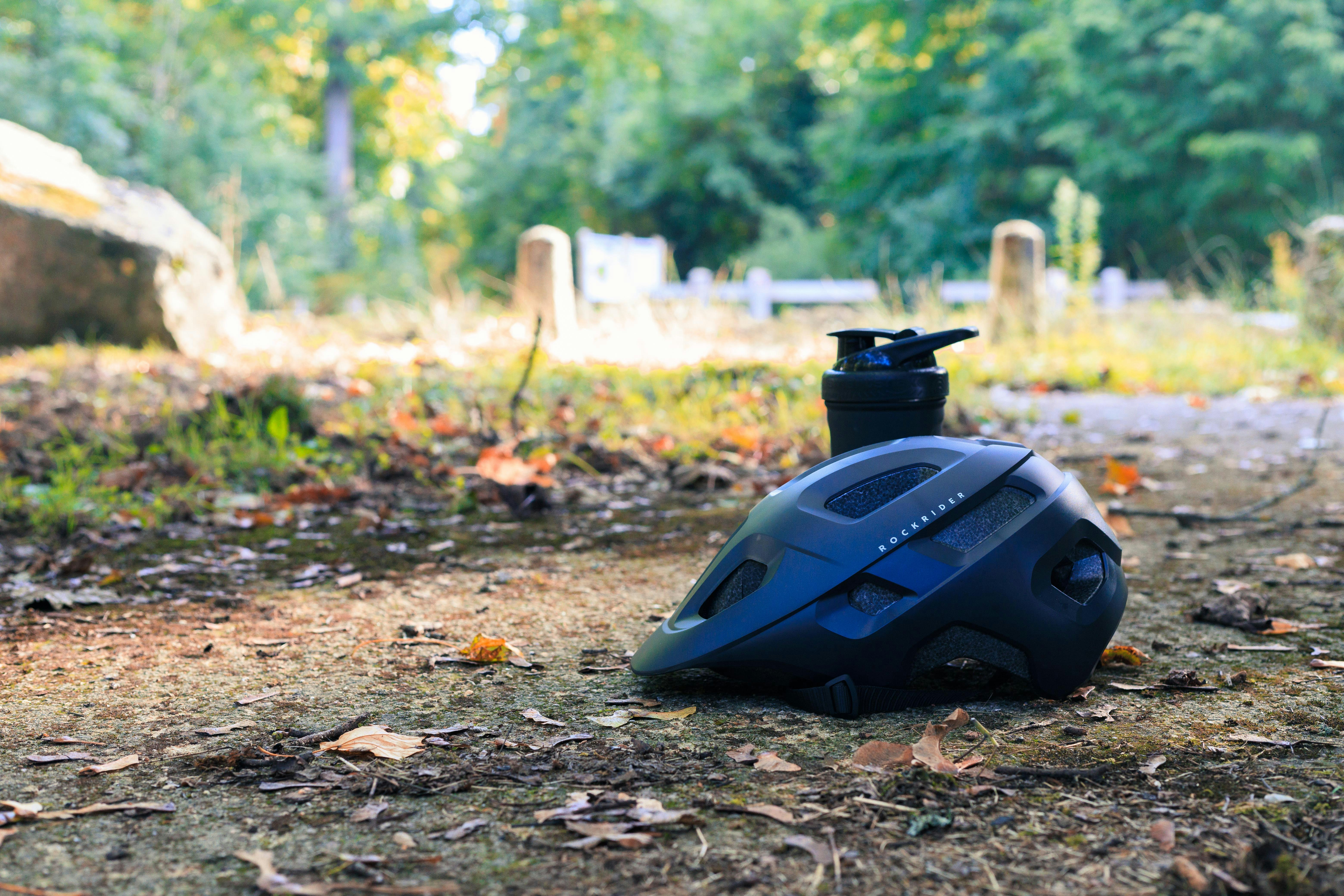 A black bike helmet rests on the ground surrounded by fallen leaves and soft sunlight filtering through trees. A water bottle sits atop the helmet, suggesting a break in an outdoor cycling adventure.