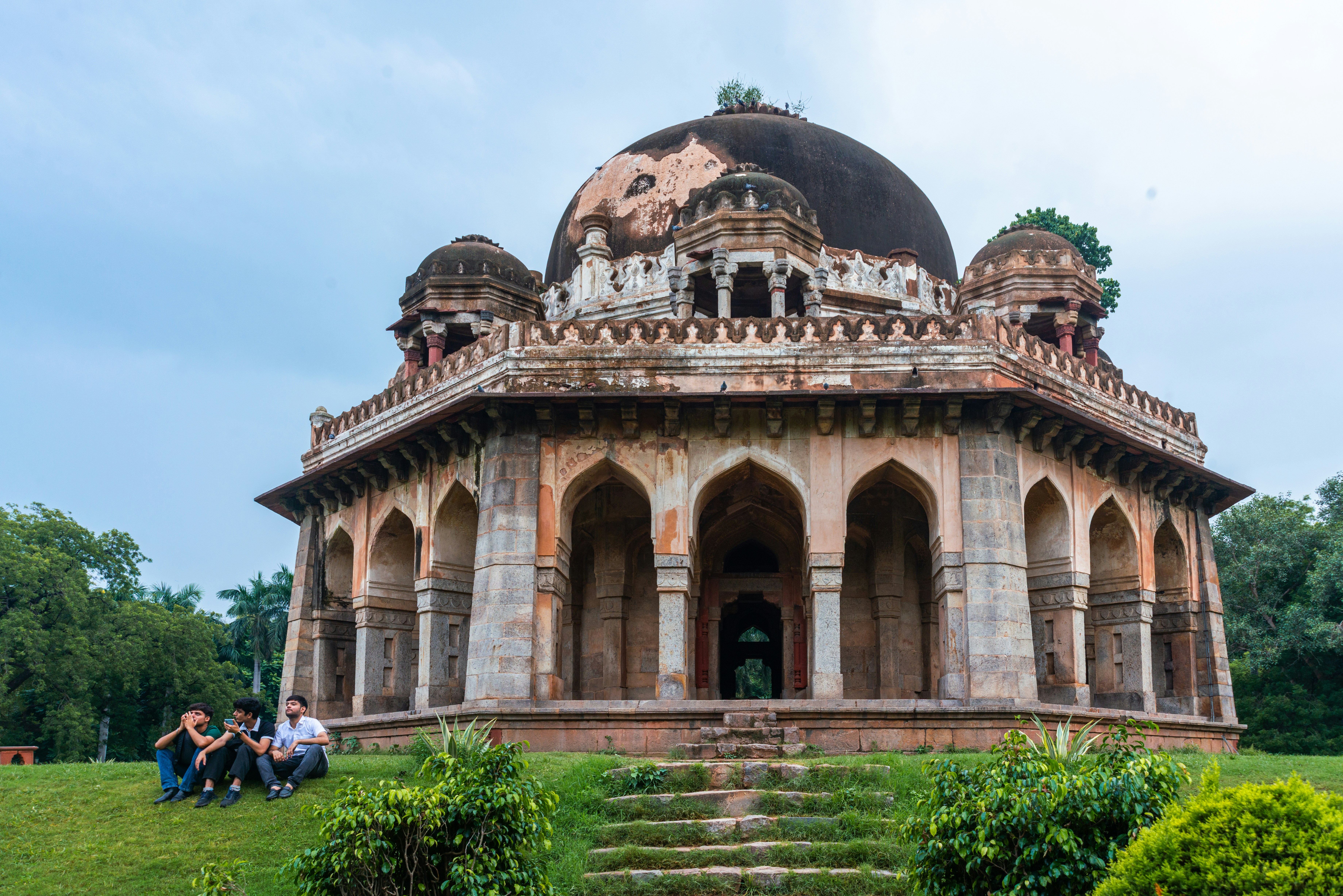 Historical mausoleum surrounded by lush greenery, with a group of friends seated on the steps, enjoying their time together.