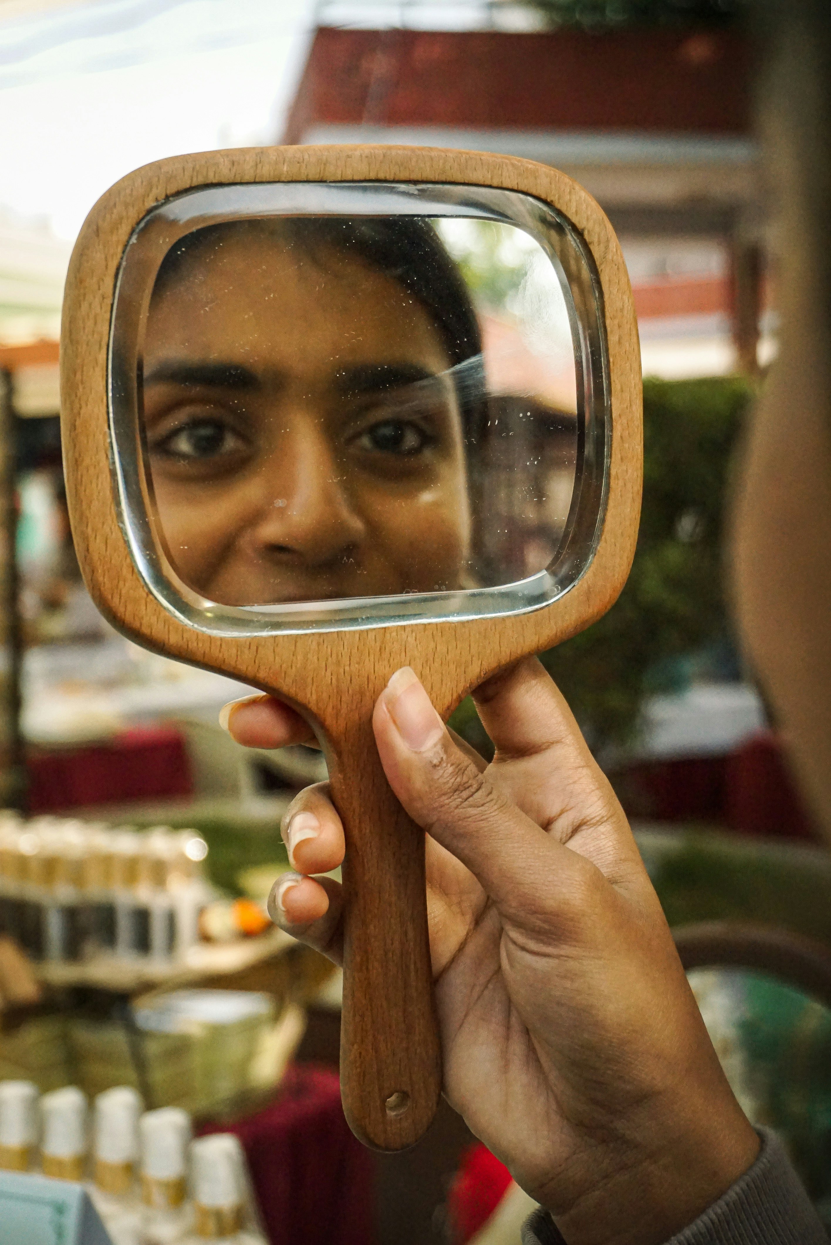 A person gazes into a handheld mirror, revealing their thoughtful expression amidst a vibrant market backdrop. The scene captures the essence of self-reflection and connection to surroundings.