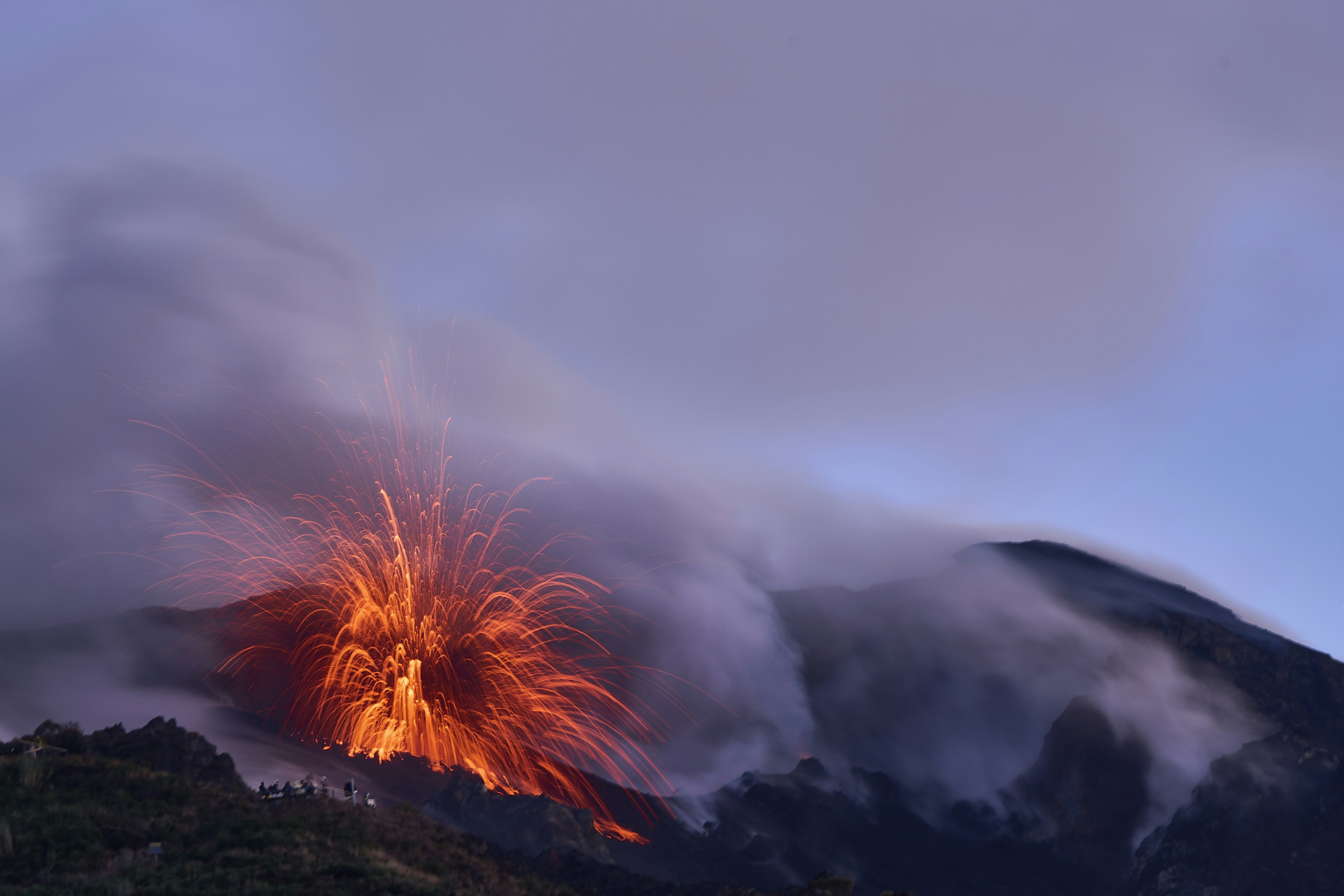 Volcanic eruption spewing vibrant lava and ash against a twilight sky, surrounded by rolling clouds and mountain silhouettes.