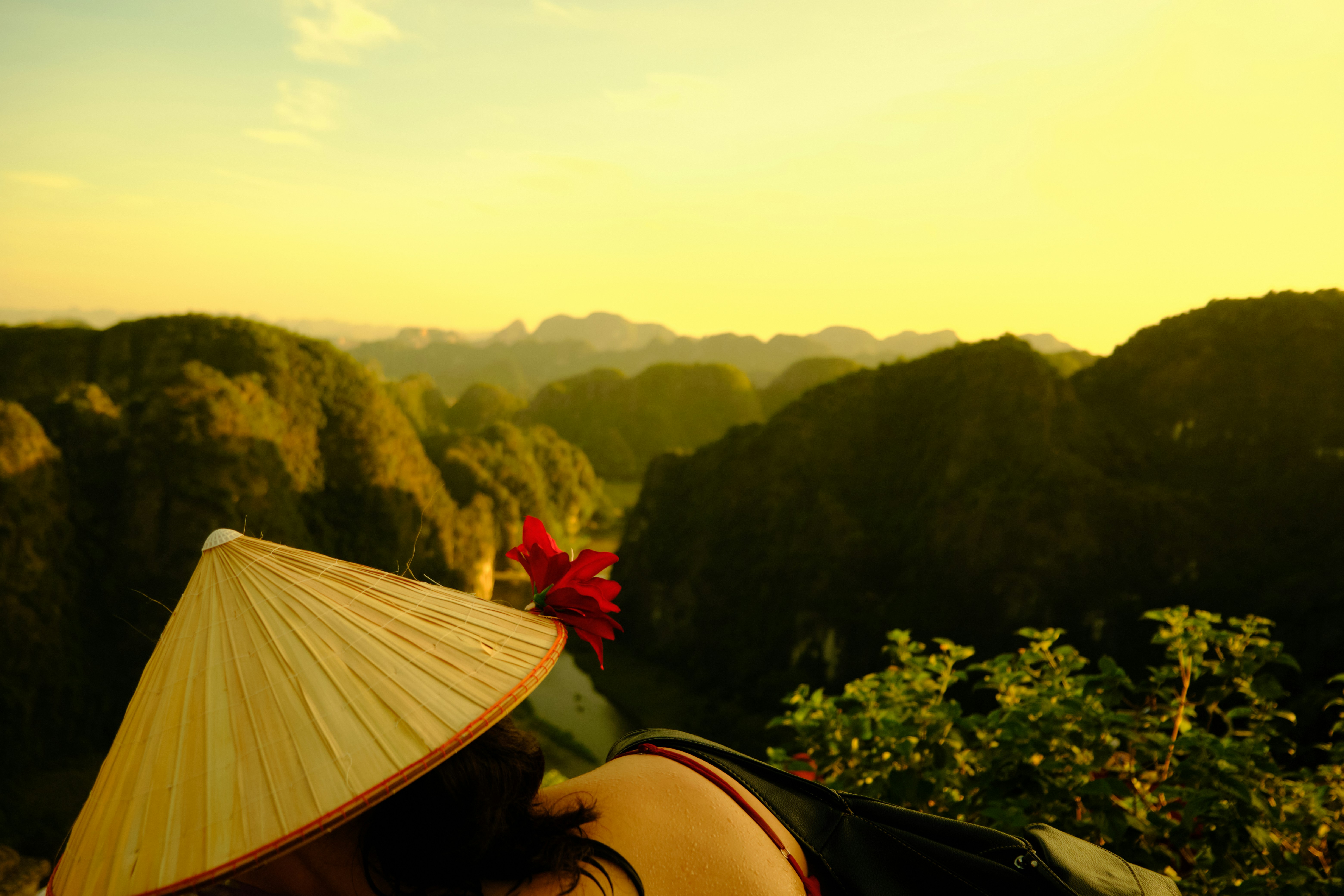 A woman wearing a conical hat gazes over lush mountainous terrain bathed in golden light during sunset.