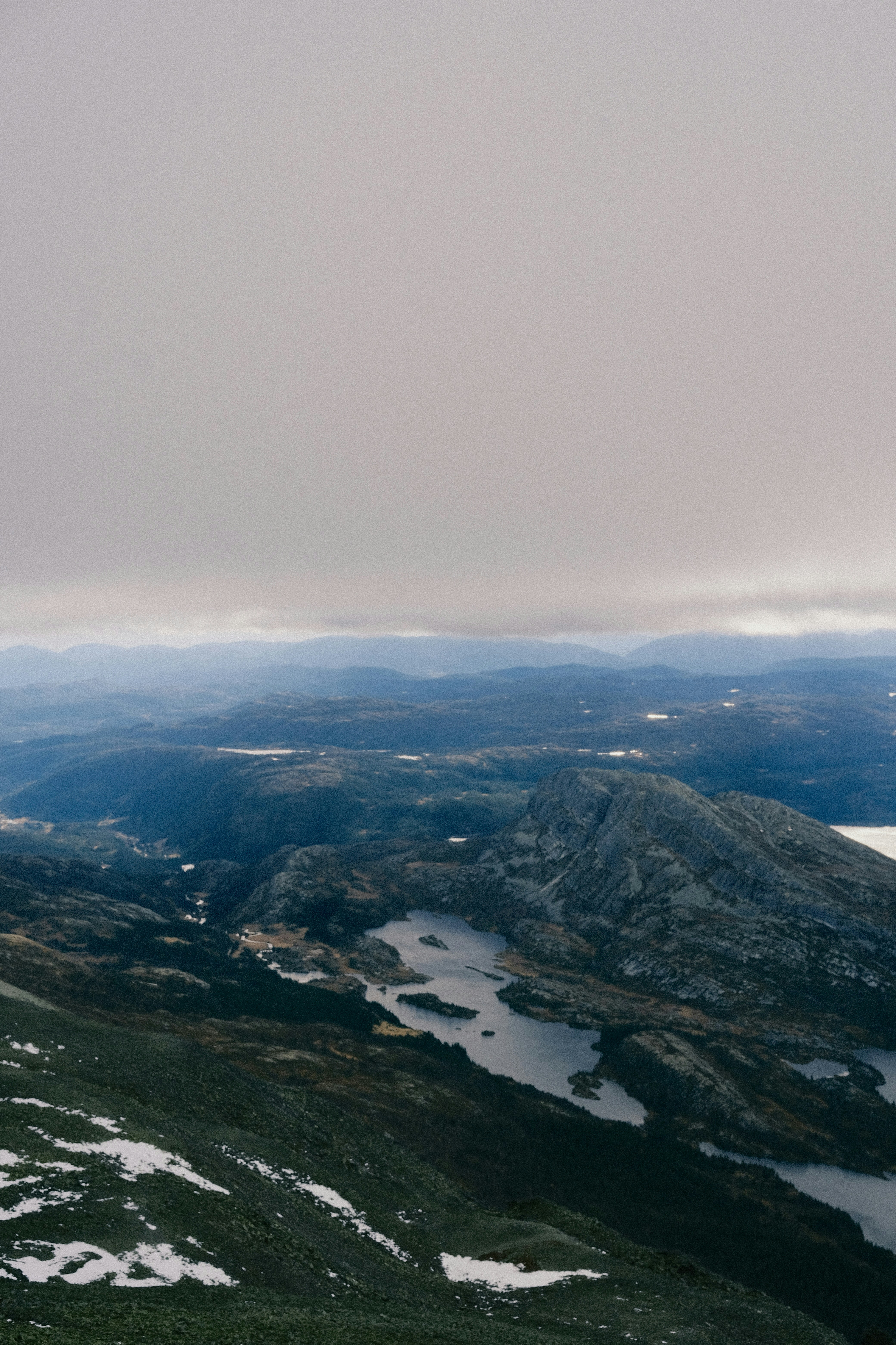 A panoramic view of a rugged mountain landscape with a winding river and patches of snow, under a cloudy sky.