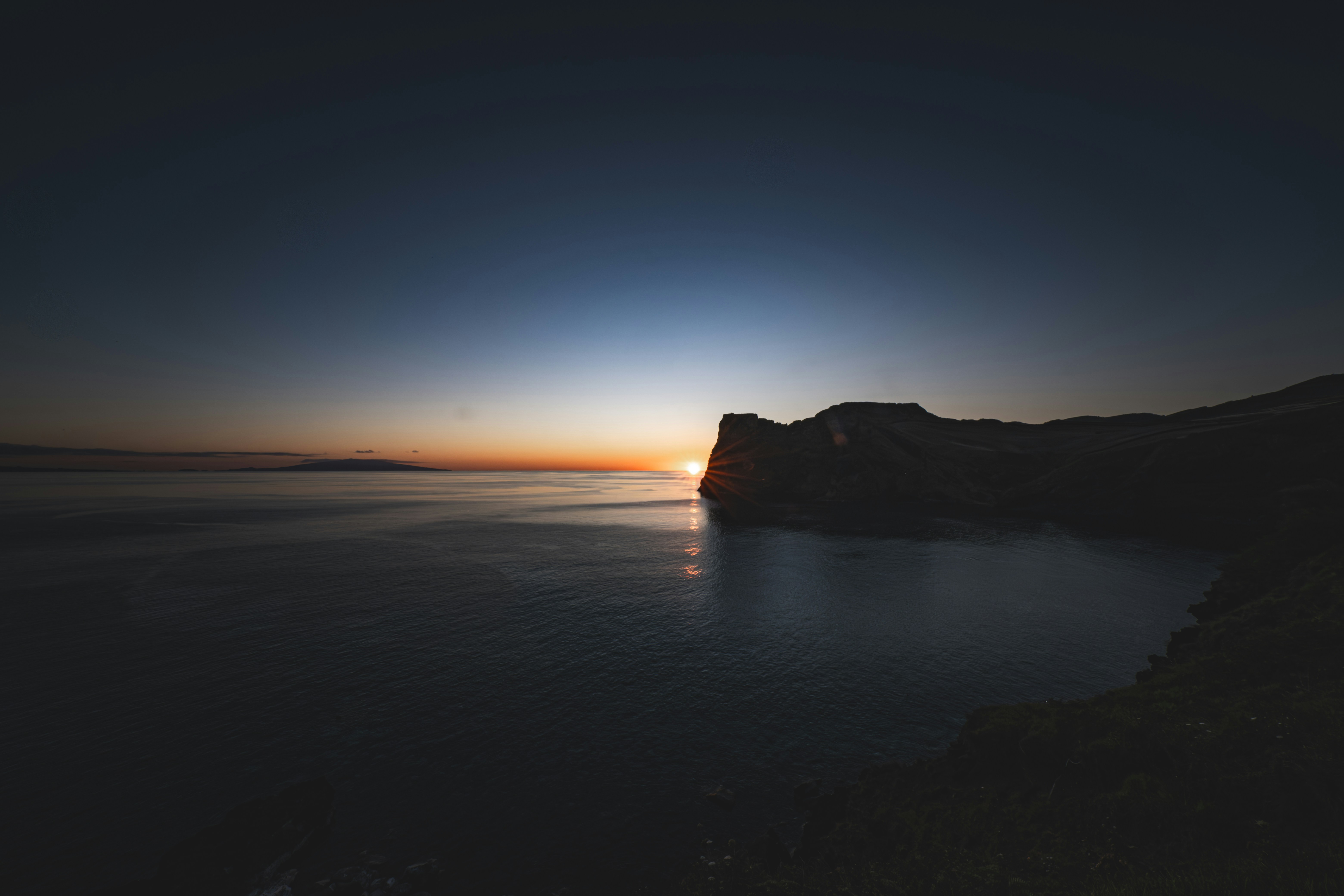Golden sunlight peeks over a rugged cliff, casting a serene glow on the calm waters below. The transition from day to night paints the sky in deep blues and warm hues.