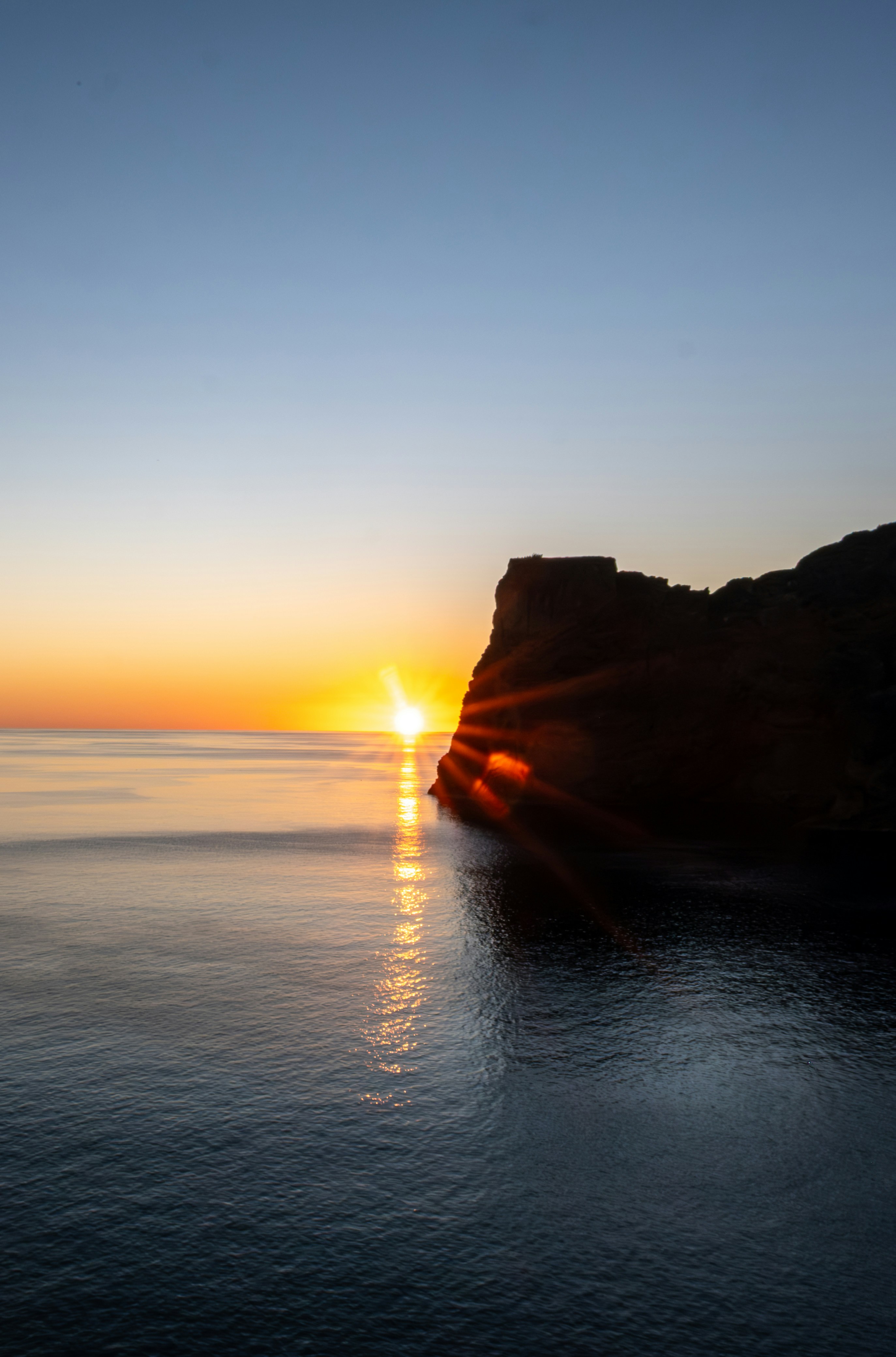 Sunset over the ocean behind a rocky cliff.