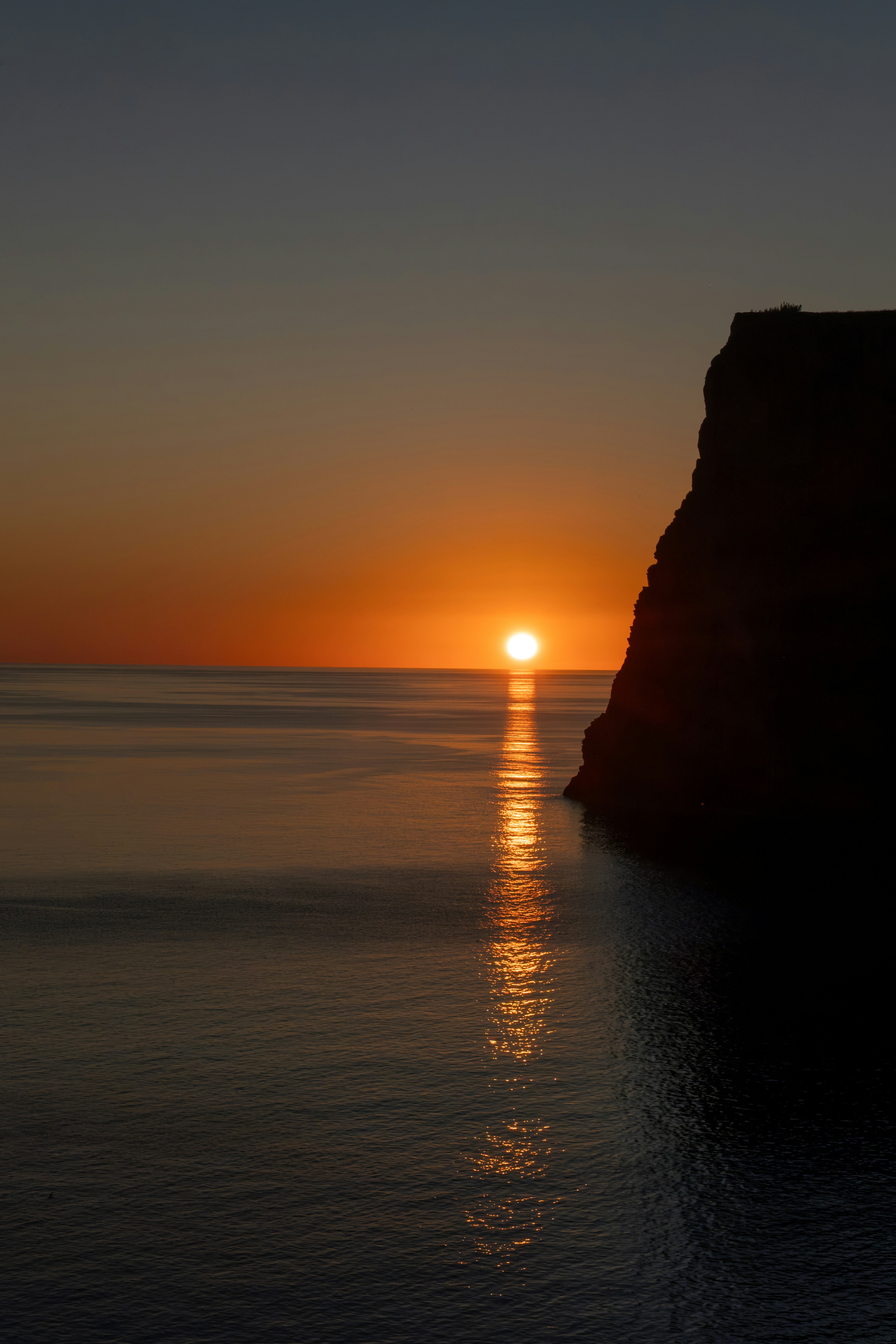 Golden sun setting behind a rugged cliff, casting a shimmering reflection on the calm sea.