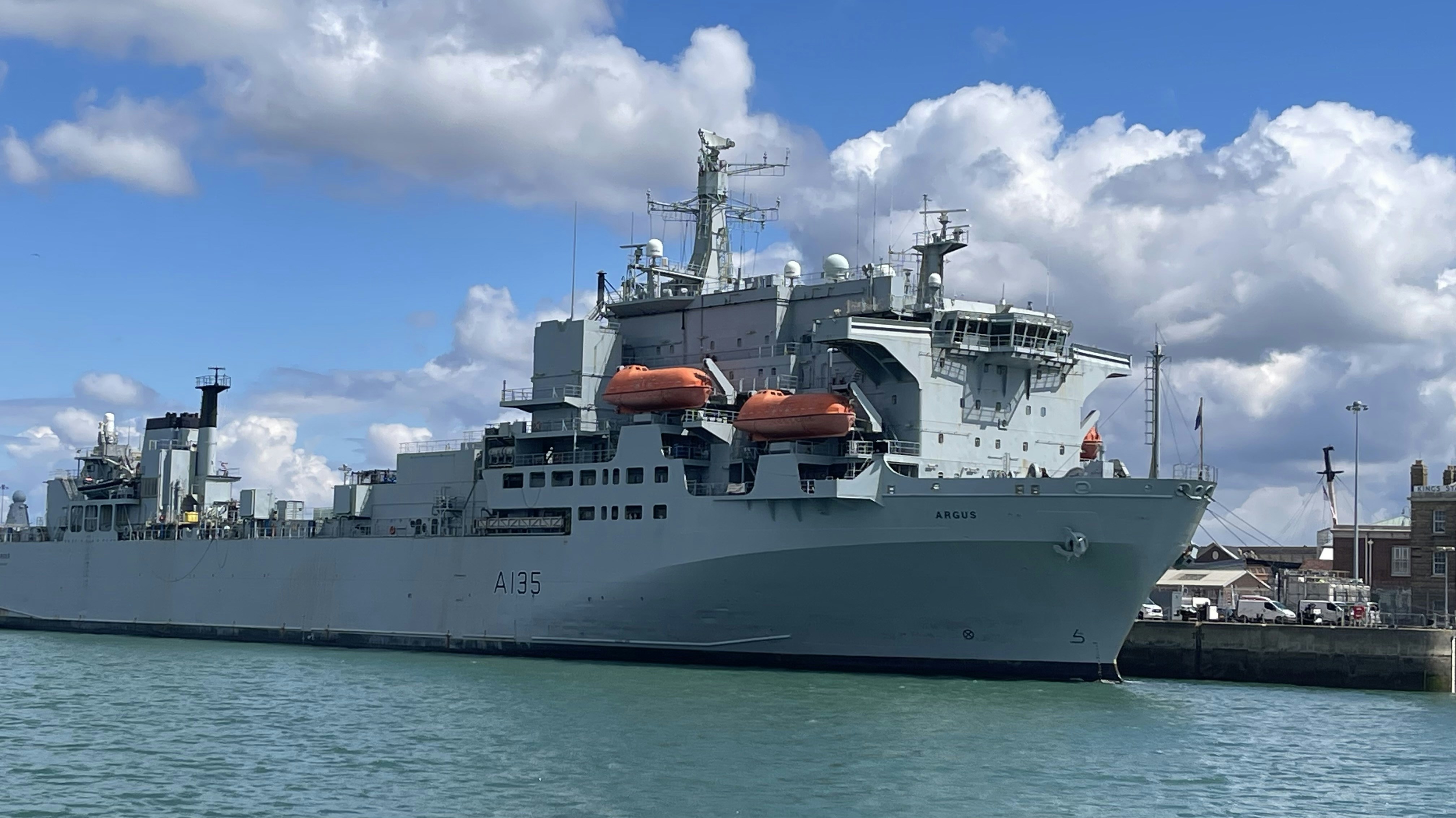 Large naval ship docked at a harbor under a partly cloudy sky, showcasing its impressive structure and lifeboats.