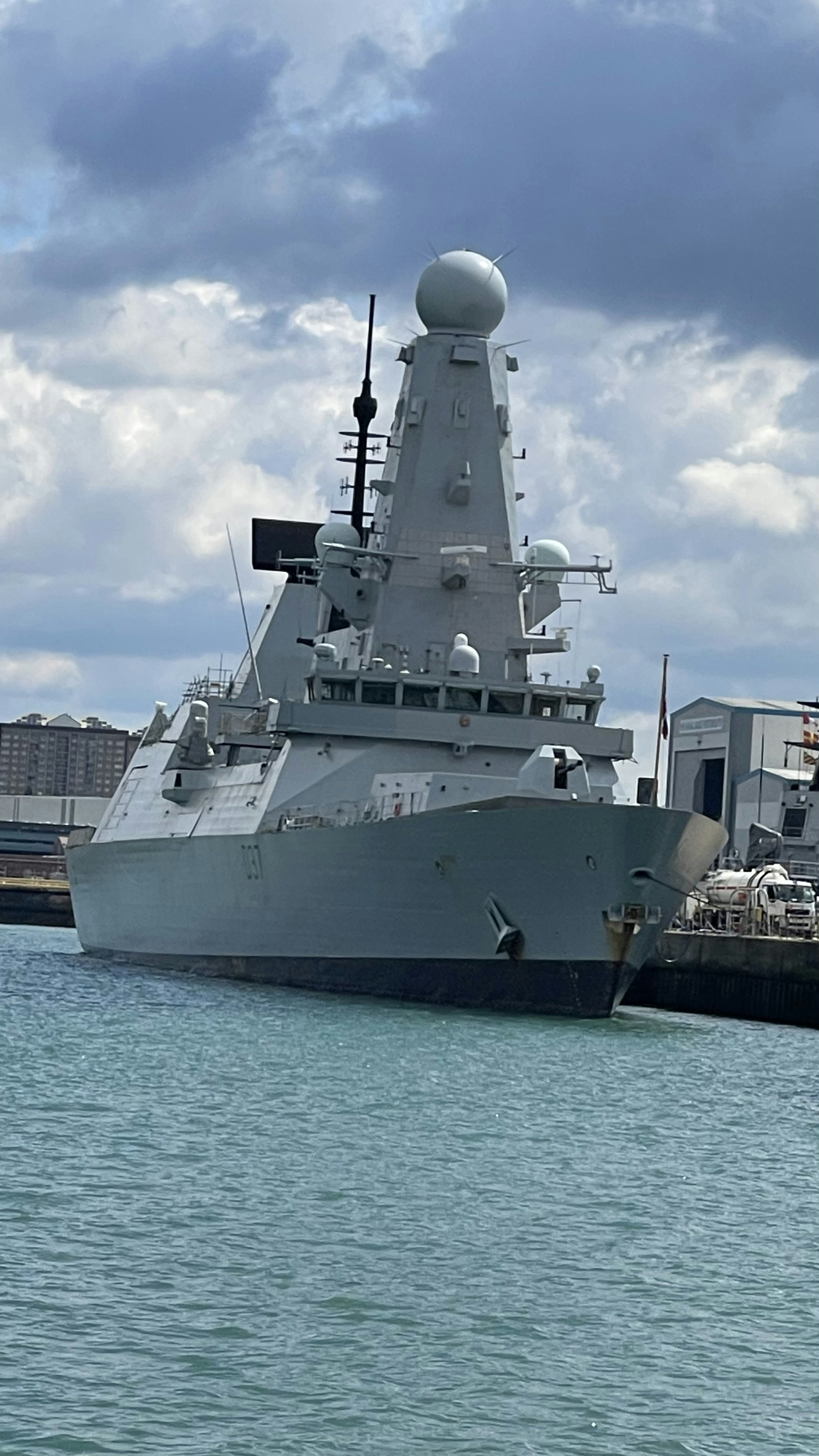 A naval destroyer docked in a harbor, showcasing advanced military design and technology against a backdrop of clouds. The vessel's sleek structure and radar systems are prominently displayed.
