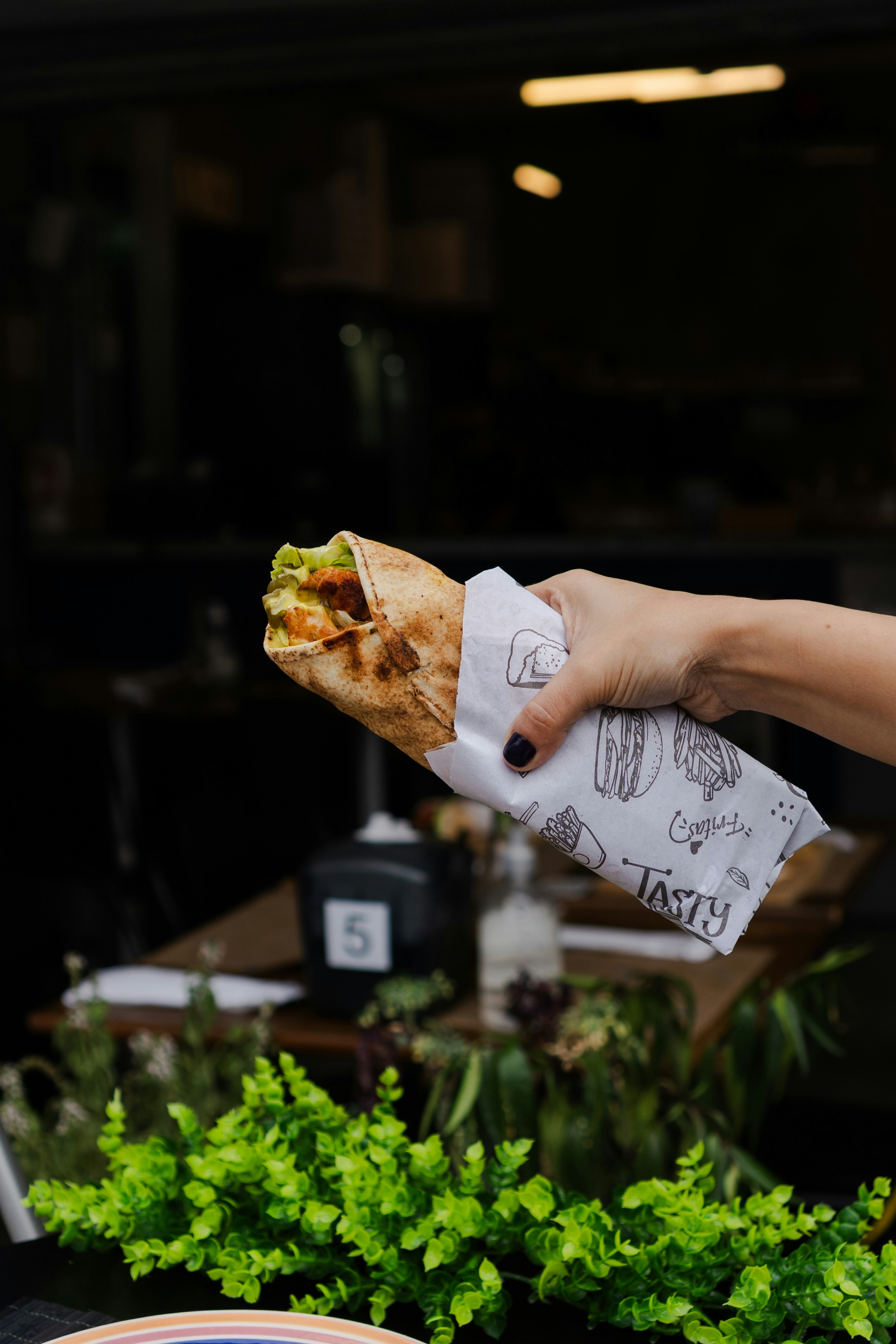 A hand holding a freshly wrapped burrito, showcasing vibrant ingredients and a decorative paper wrap. Lush greenery and a rustic table setting enhance the scene.