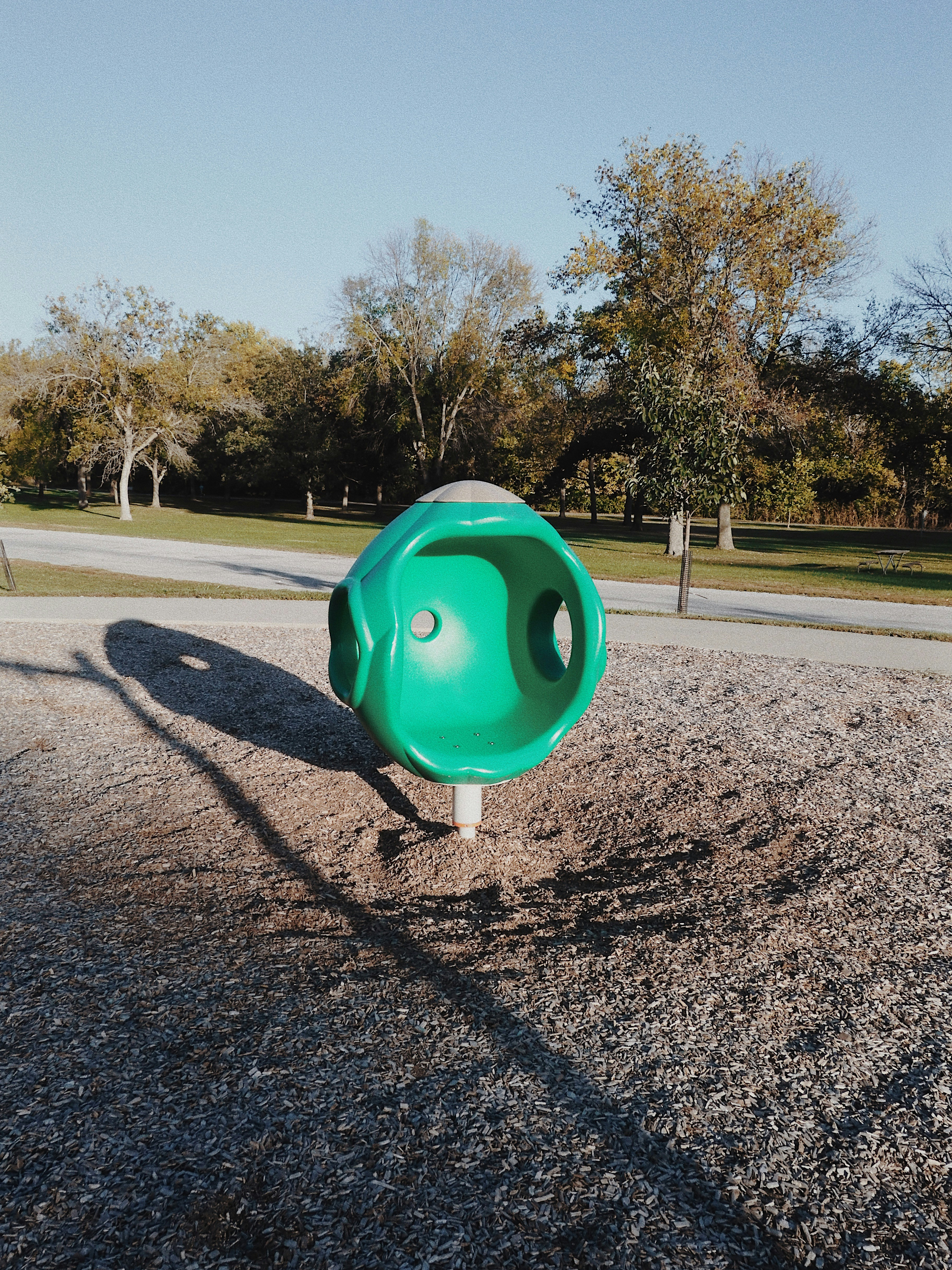 Taken in Iowa City. A modern green play structure stands out against autumn trees and mulch. The abstract design invites imaginative play, framed by seasonal colors and quiet park surroundings. | A green playground spinner casts a long shadow.