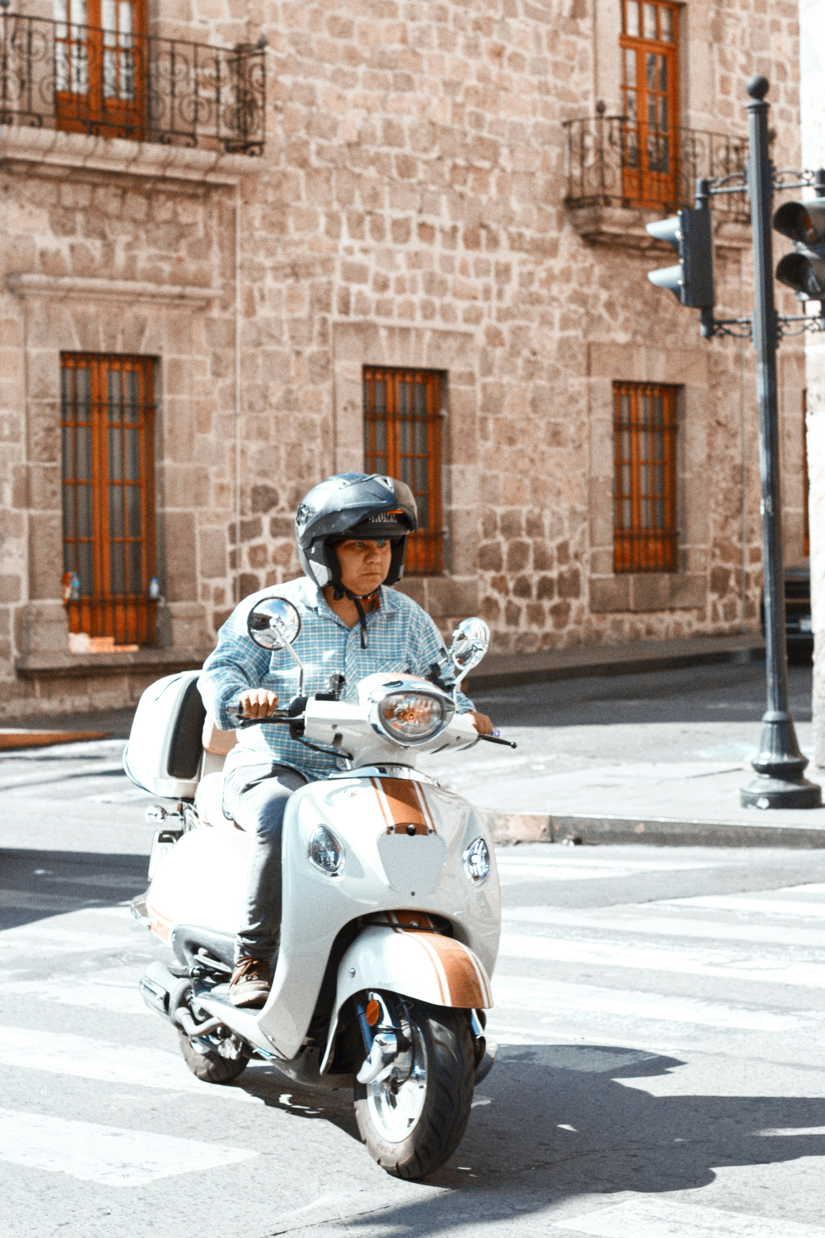 A rider on a scooter navigates a quaint, sunlit street lined with historic stone buildings and wooden windows.