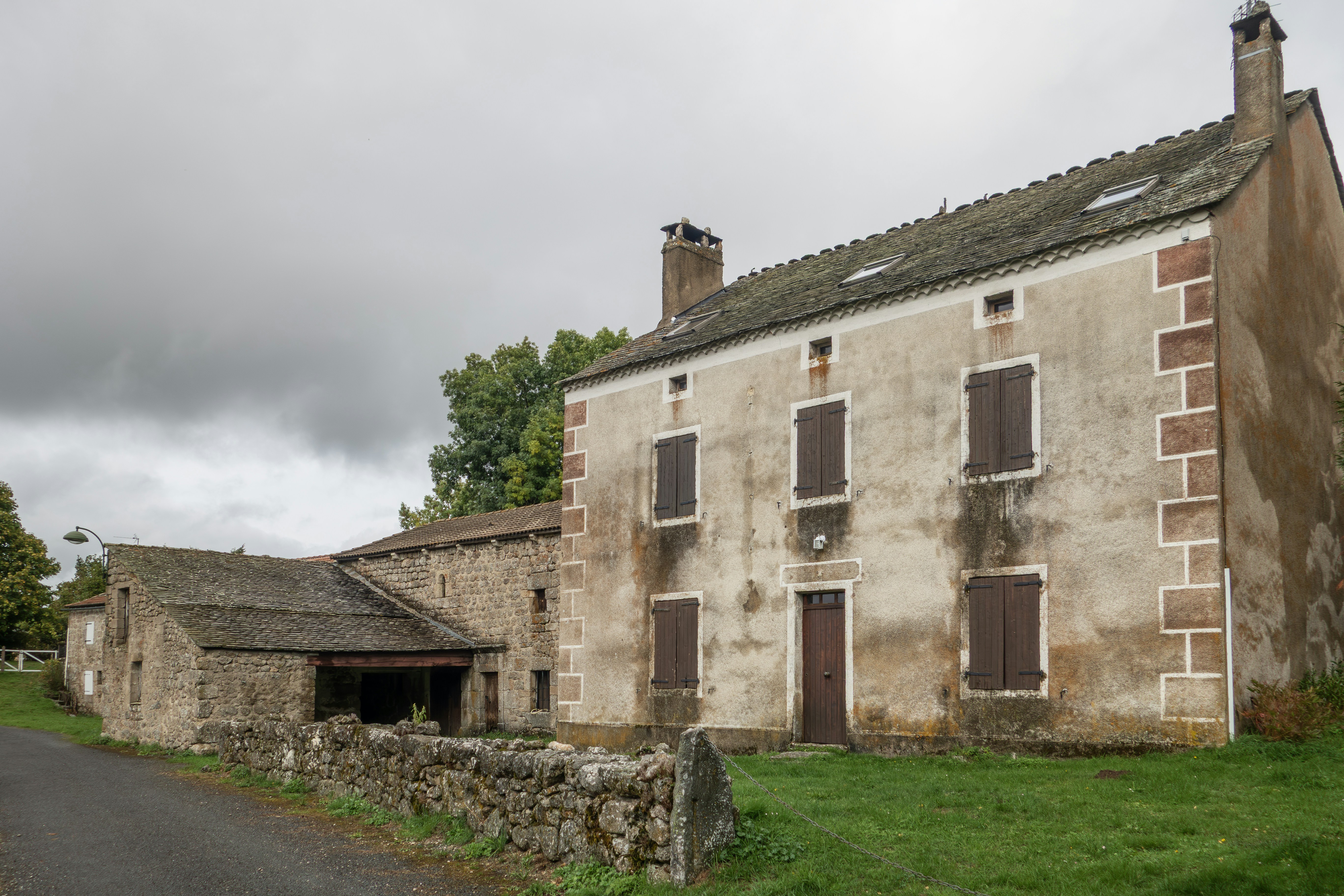 Sur ce chemin, l’homme ne conquiert rien. Il écoute. Il marche entre les pierres et les nuages, là où Stevenson jadis chercha le silence et la vérité. Chaque pas est une mémoire, chaque paysage une page ouverte. Ce n’est pas la France qu’on traverse, mais un monde en miniature — fait de lumière, de solitude, et d rencontres. | Old stone farmhouse with shutters under cloudy sky