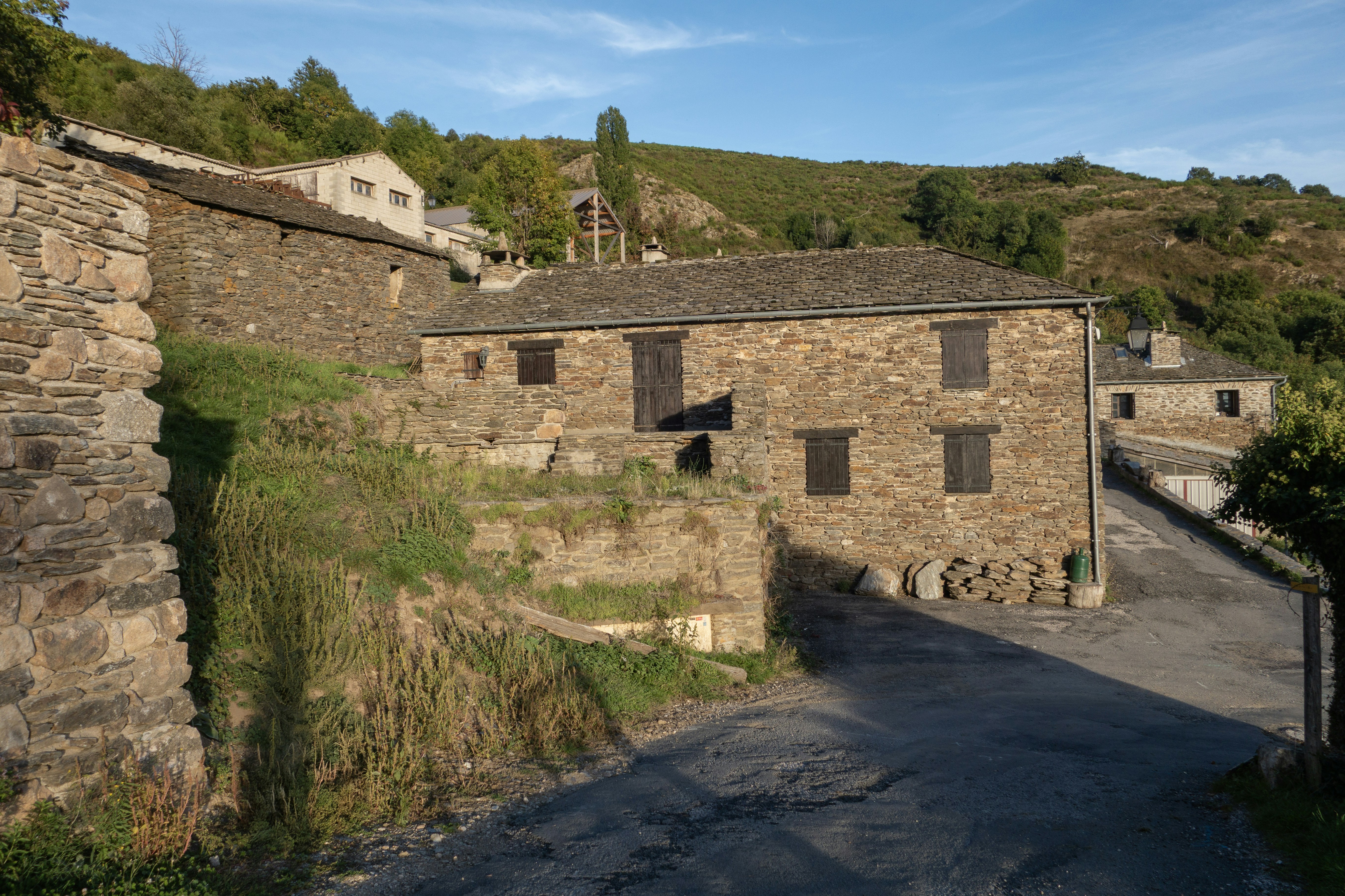 Rustic stone houses nestled in a hillside, surrounded by lush greenery and a winding path. The scene captures the tranquility of rural architecture harmonizing with the landscape.