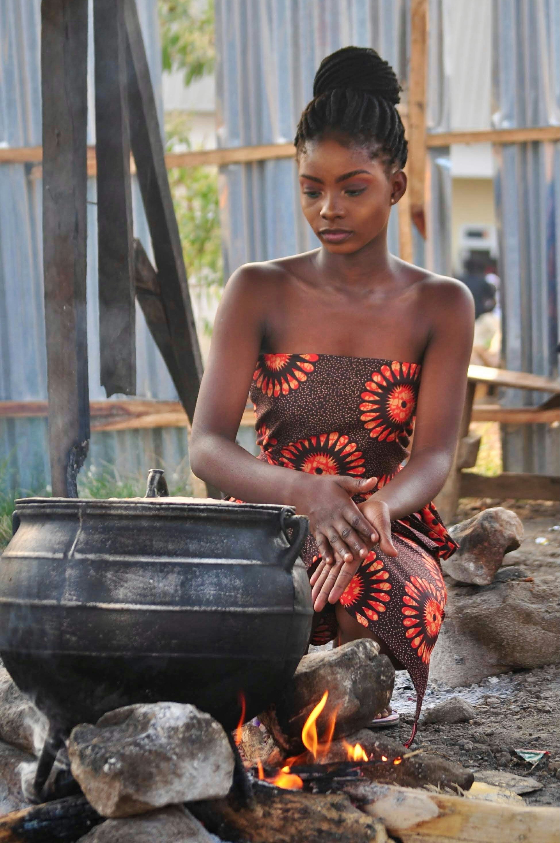A series of authentic images capturing a beautiful young Nigerian woman in a rural setting. Dressed in vibrant traditional Ankara print fabric with her hair in a braided bun, she is seen cooking a meal in a large cast-iron pot over a traditional three-stone fire. Other images show her serene expressions and her strength as she carries a bucket on her head through a dry, grassy field. These photos celebrate African culture, heritage, resilience, and the simple joys of daily life. | Young woman cooking over an open fire