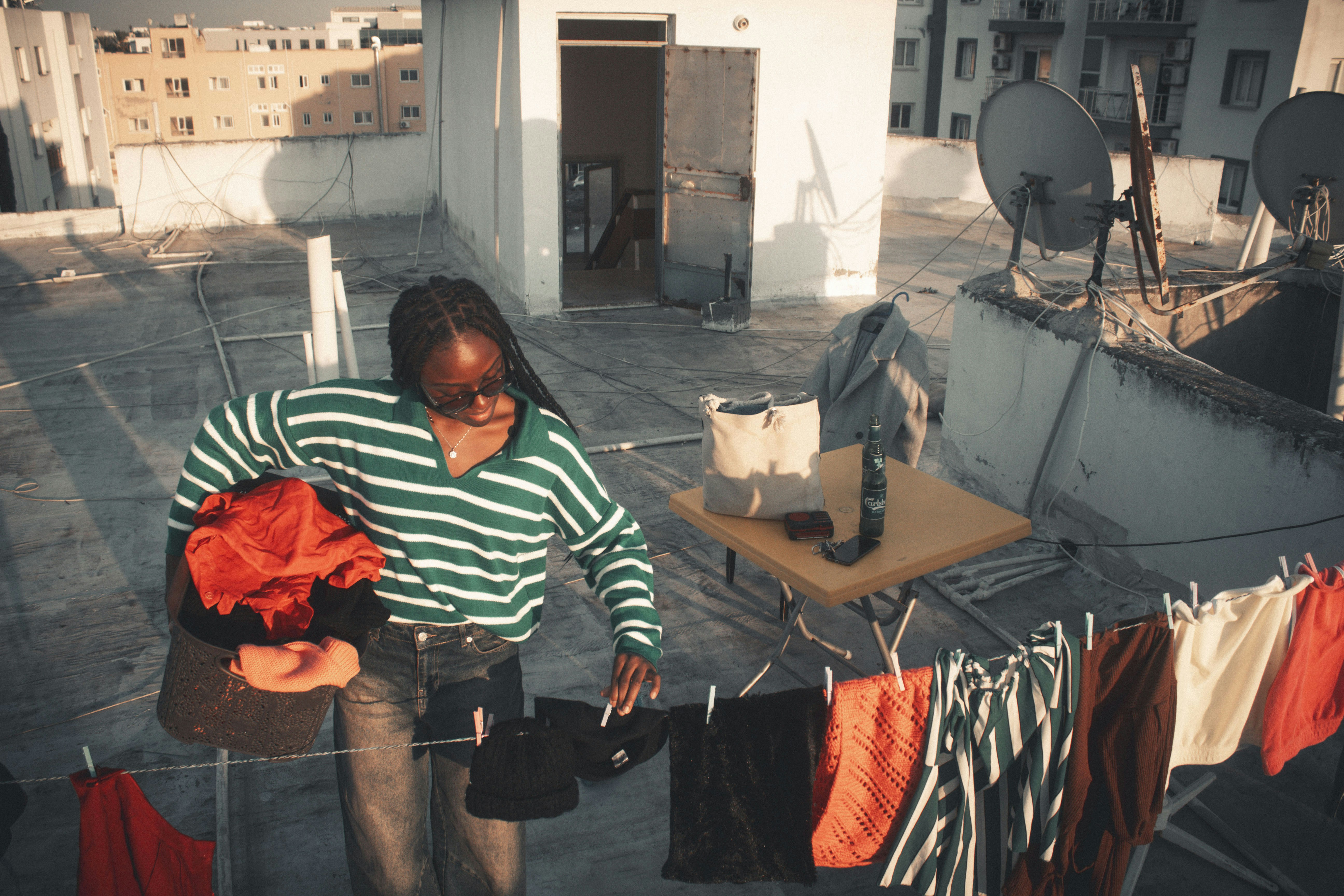 A person dressed in a striped sweater carries a basket of laundry on a rooftop, with colorful clothes hanging on a line and urban buildings in the background.