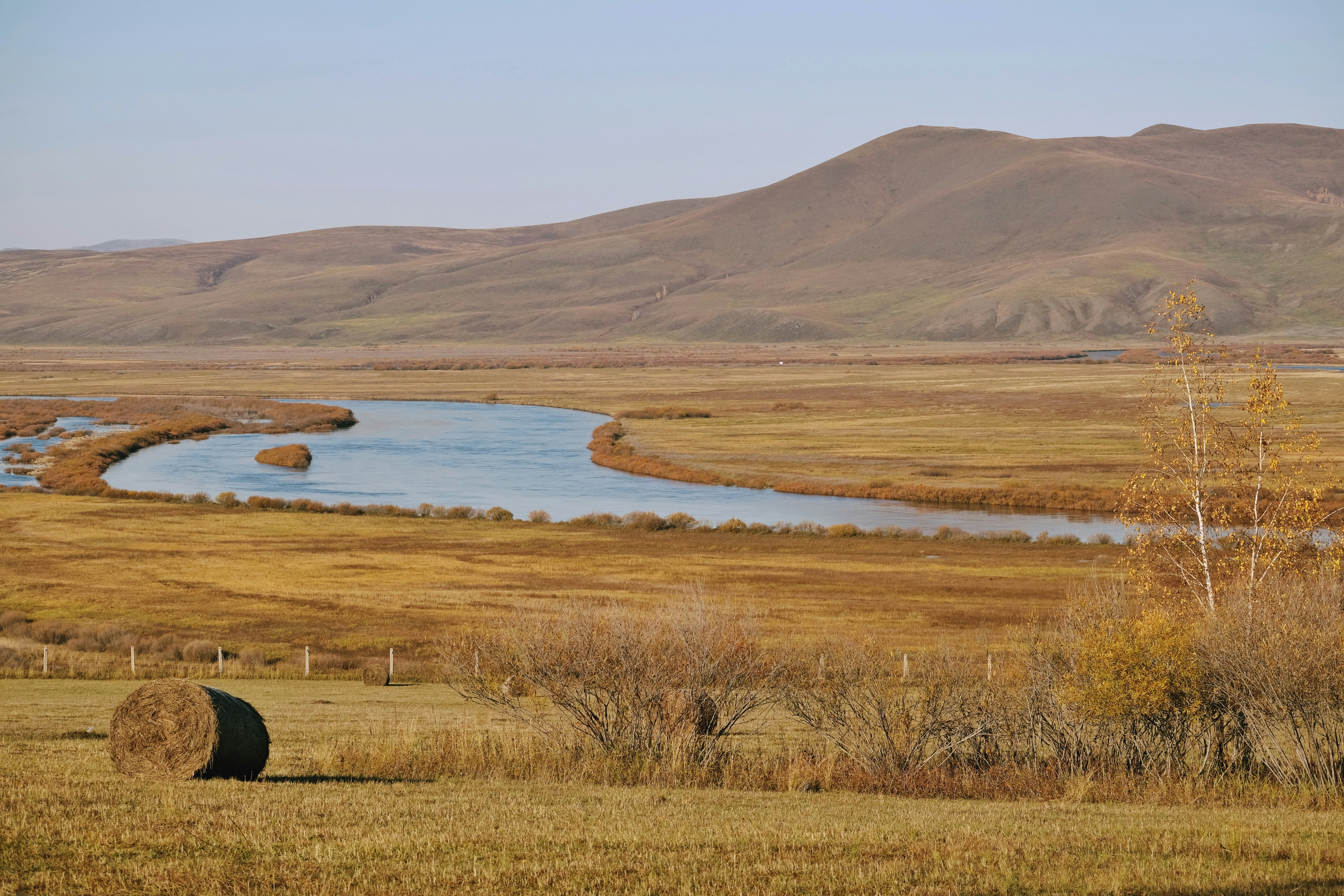 A single hay bale sits in a dry, grassy plain with a curving river and distant rolling hills. | Hay bales in a golden field with a winding river.