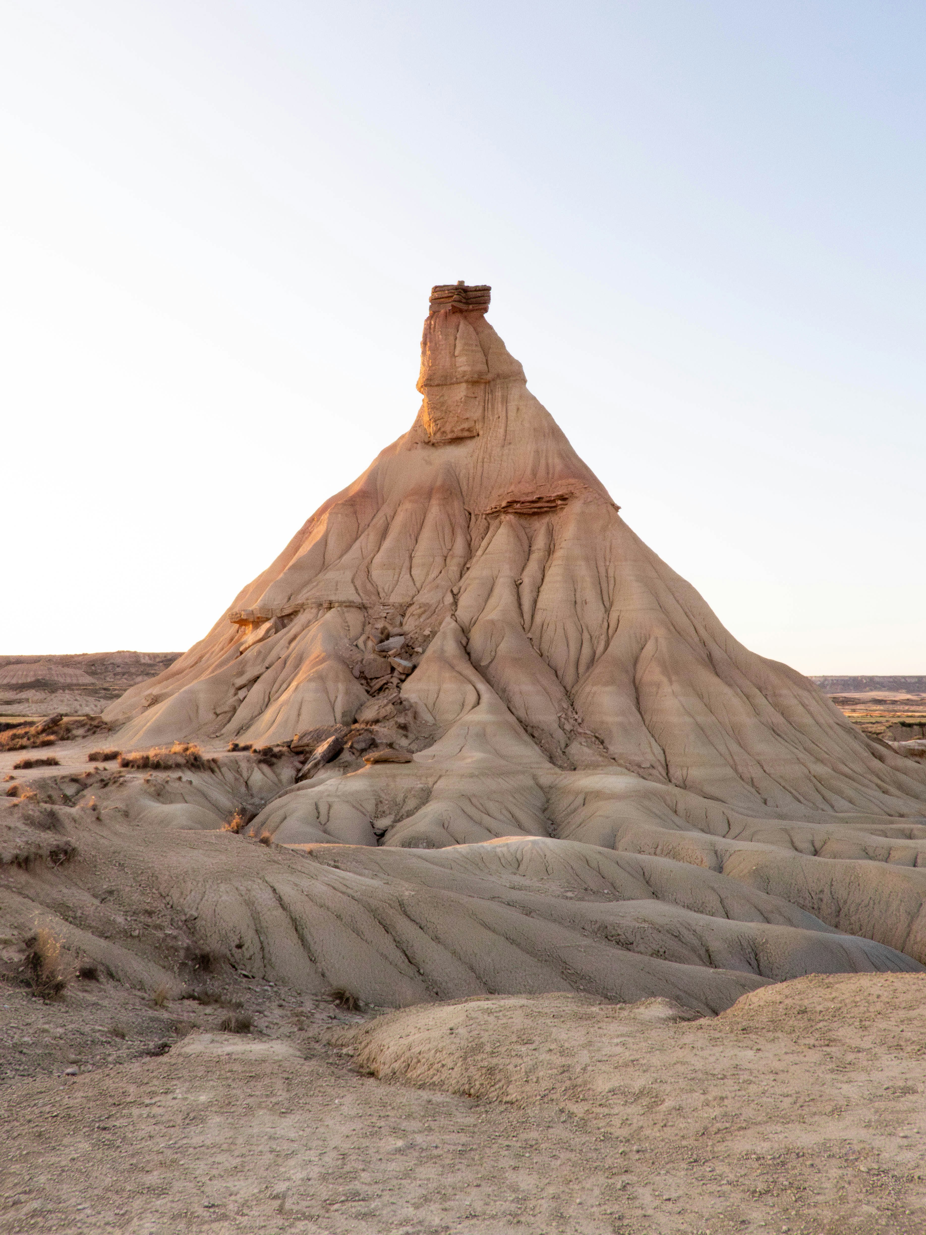 A conical rock formation rises from the eroded ground in the Bardenas Reales desert, Navarra, Spain. The structure has layered sediment and a flat rock at the top, surrounded by dry terrain. | Unique rock formation in a desert landscape at sunset