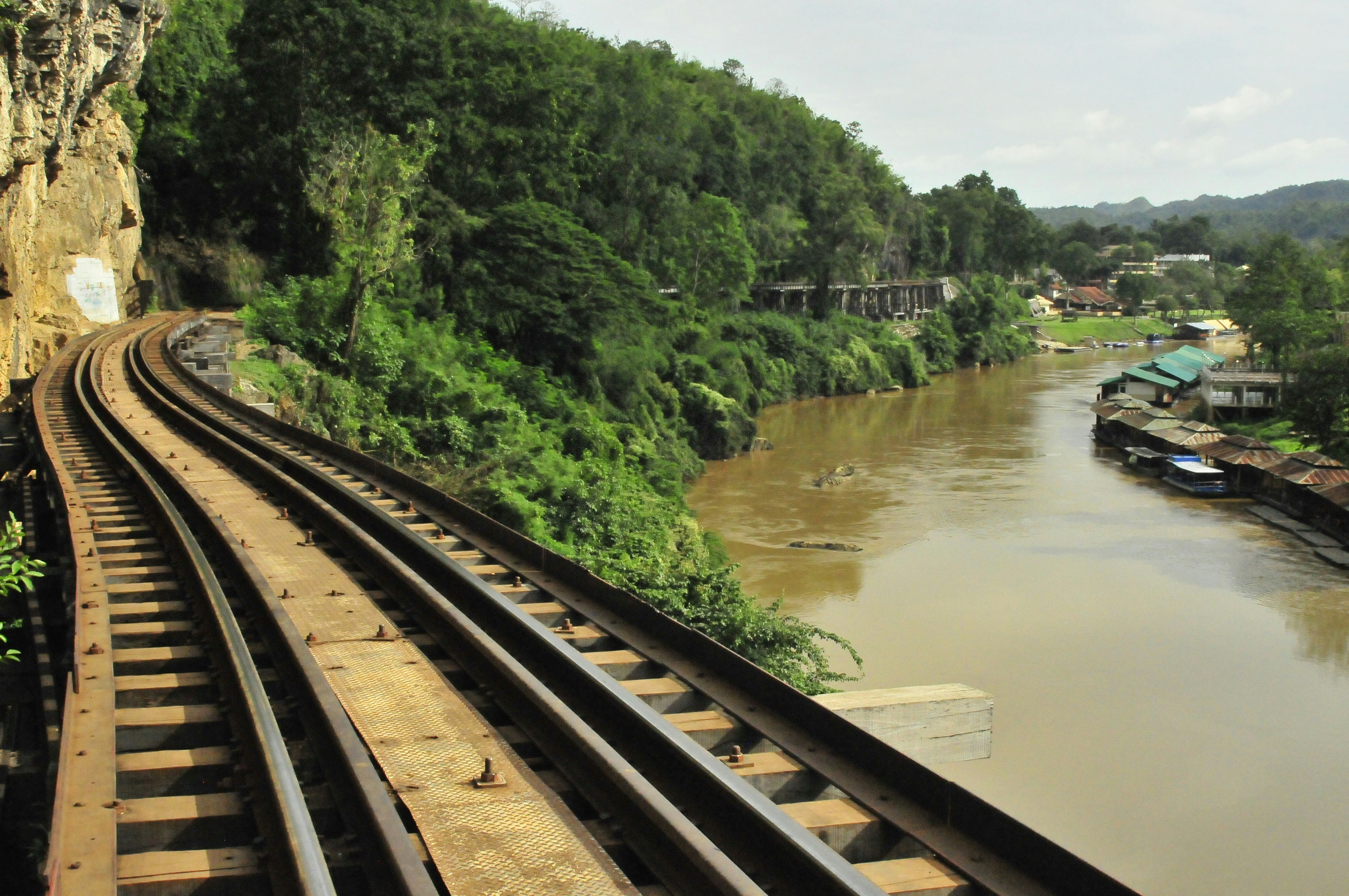 The railway next to the river Kwai | Train tracks run along a river with lush green hills.
