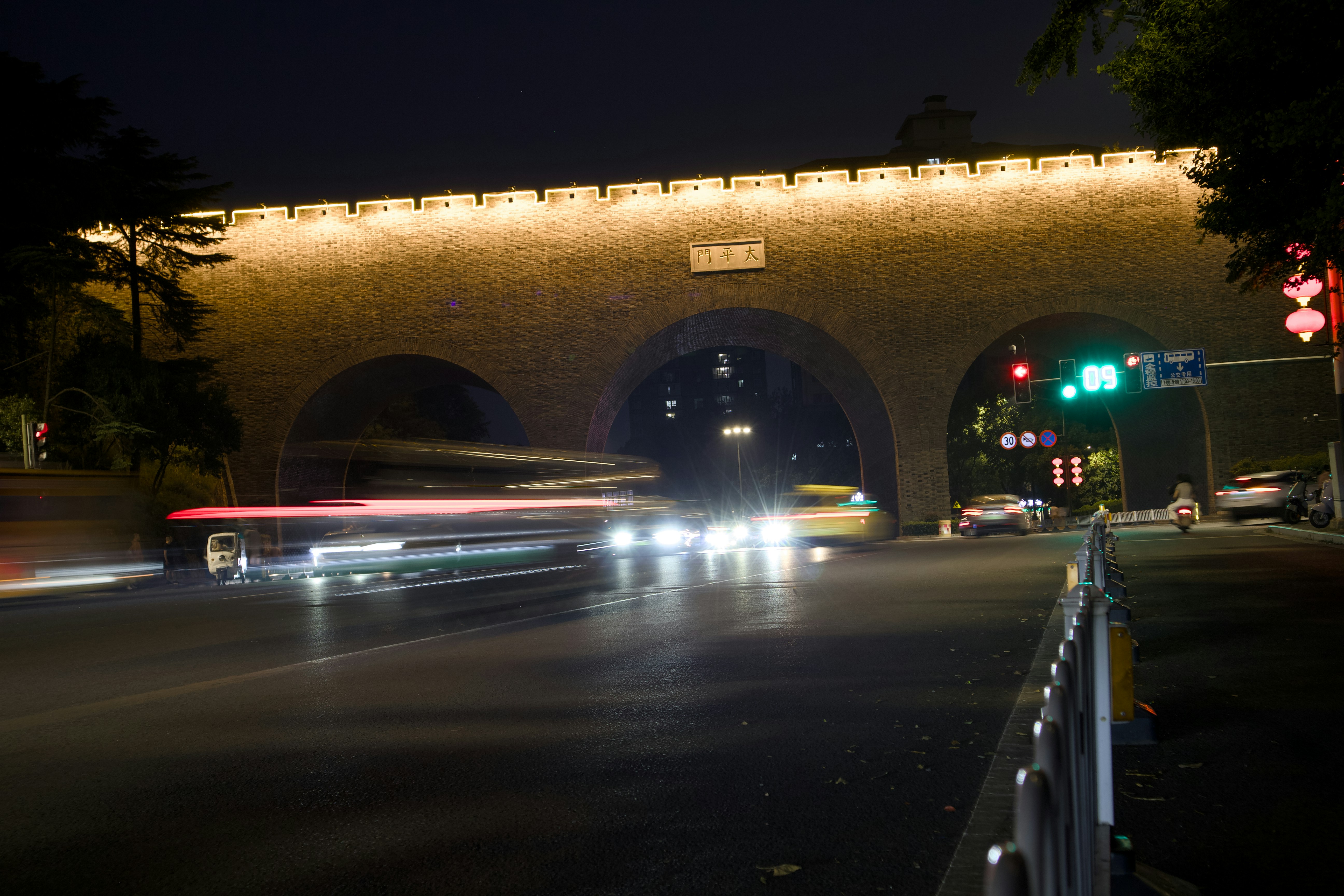 Cars driving under illuminated ancient stone archway at night.