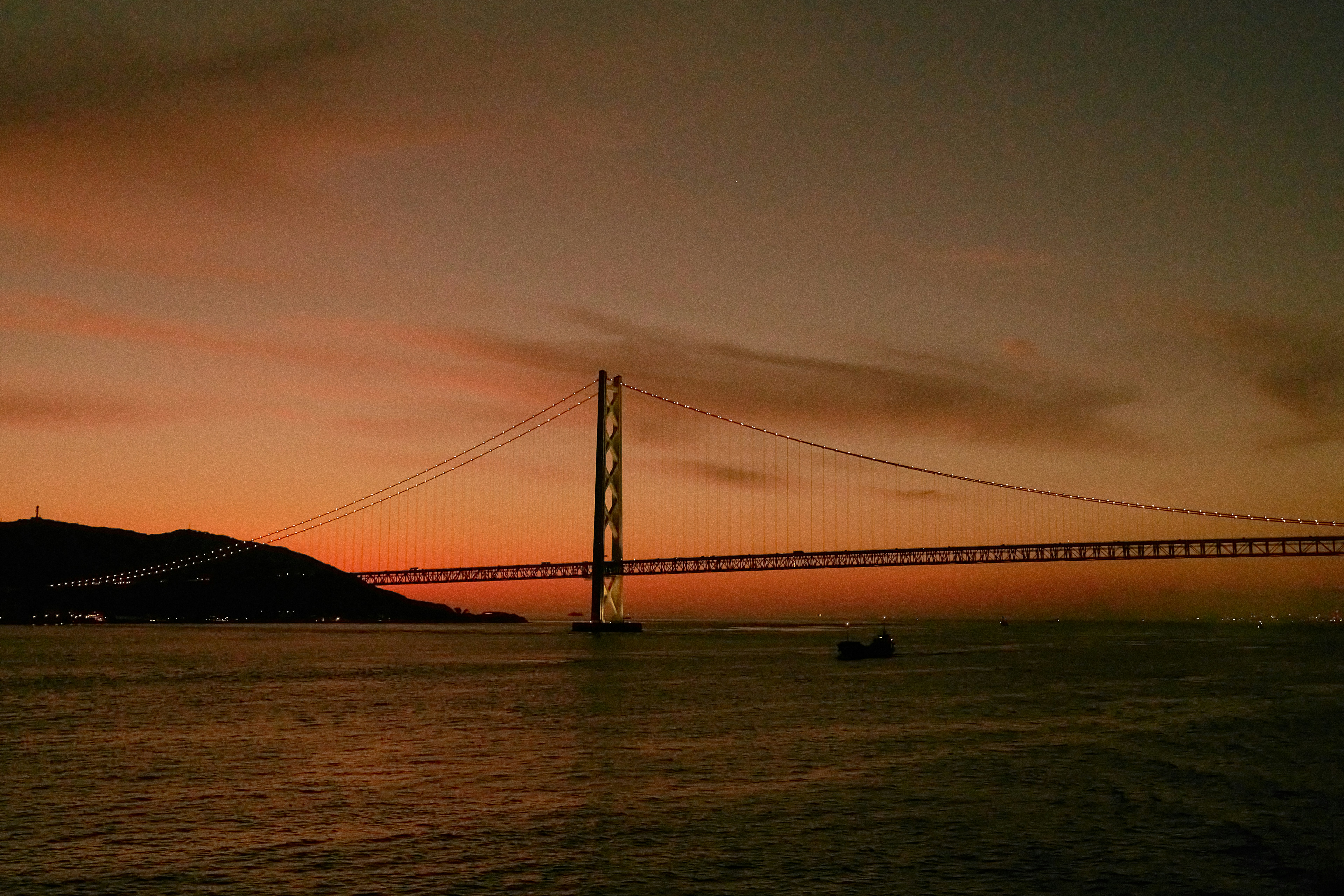 Illuminated bridge spanning a calm sea at twilight, with soft hues of orange and pink in the sky. A small boat navigates the tranquil waters beneath the structure.